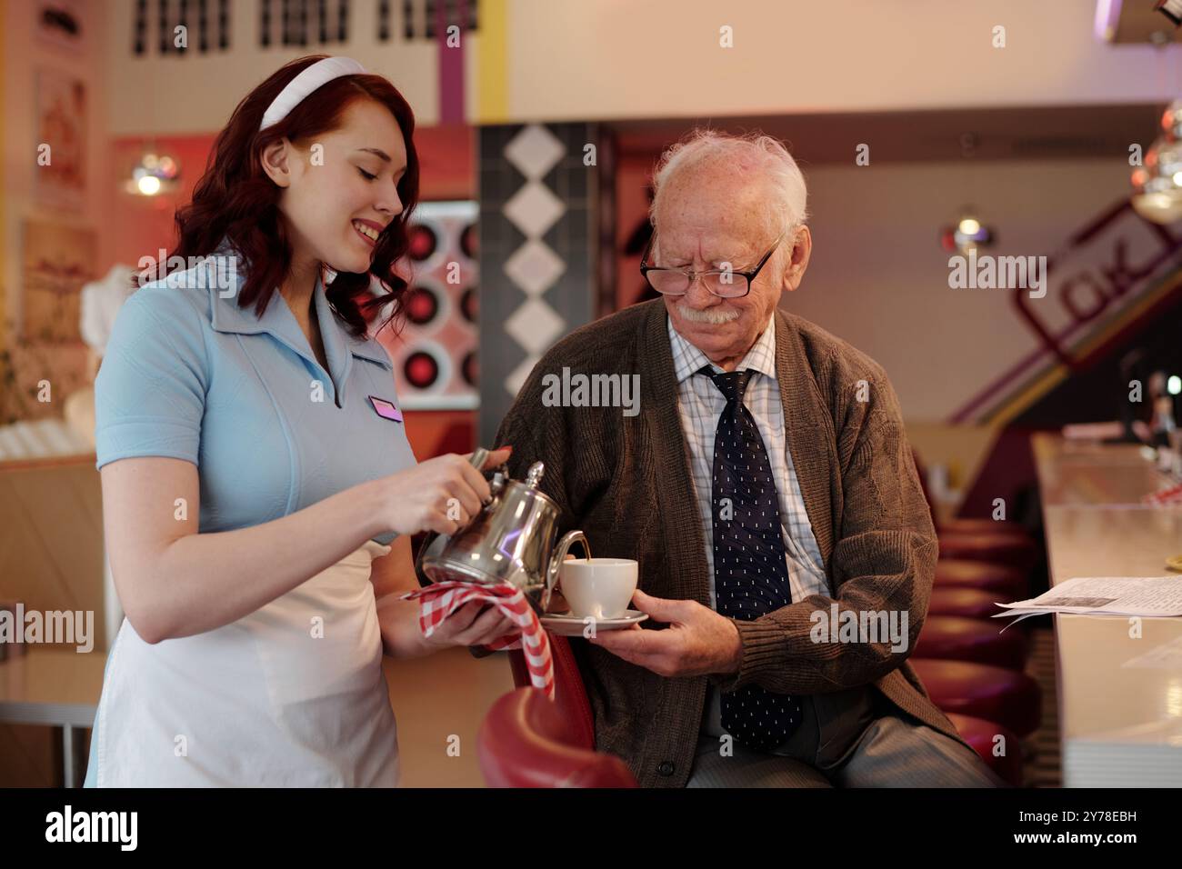 Smiling waitress serving coffee to elderly man seated at retro diner ...