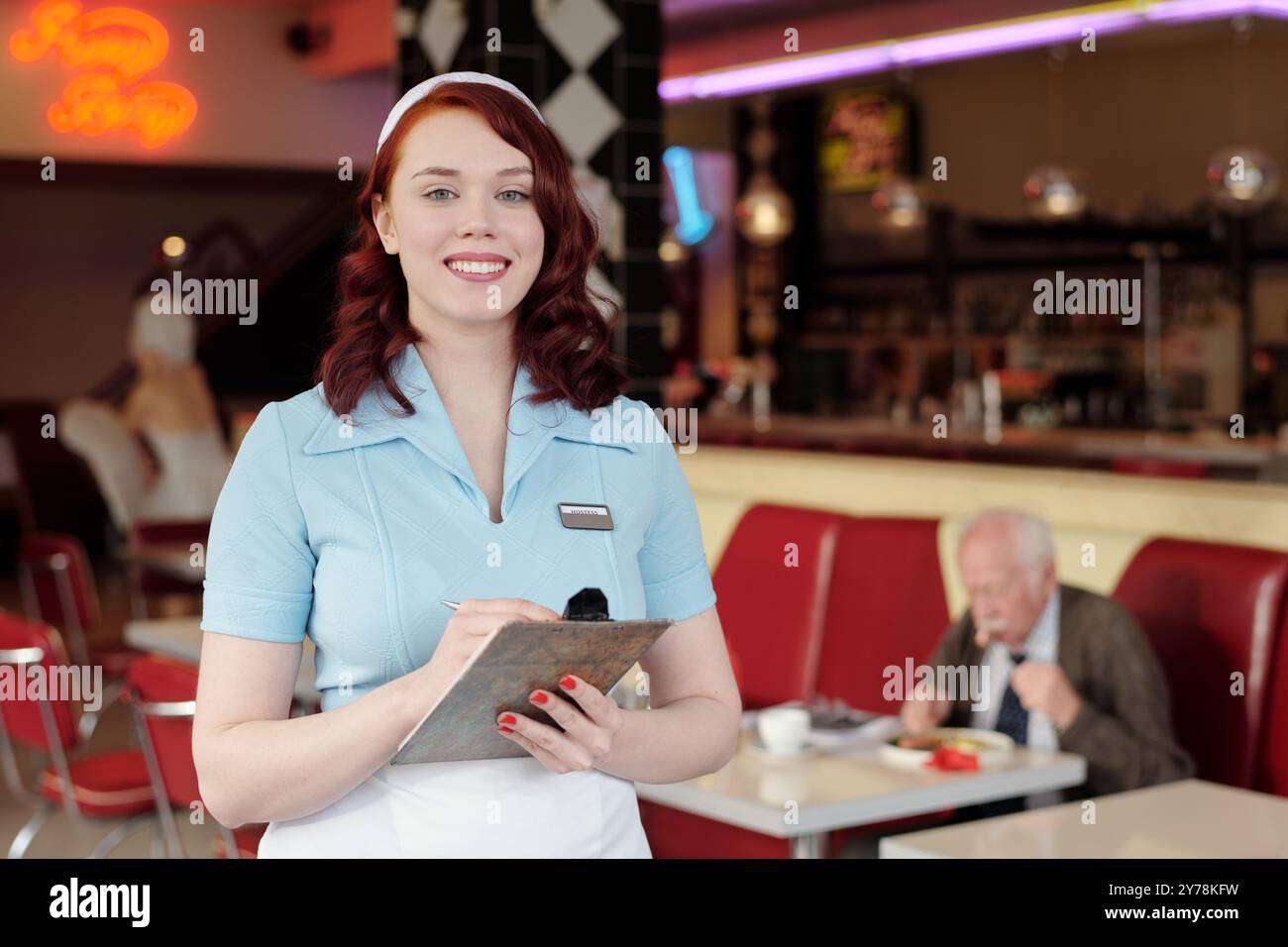 Smiling waitress holding a notepad in 1950s style diner with red upholstery and elderly man ...