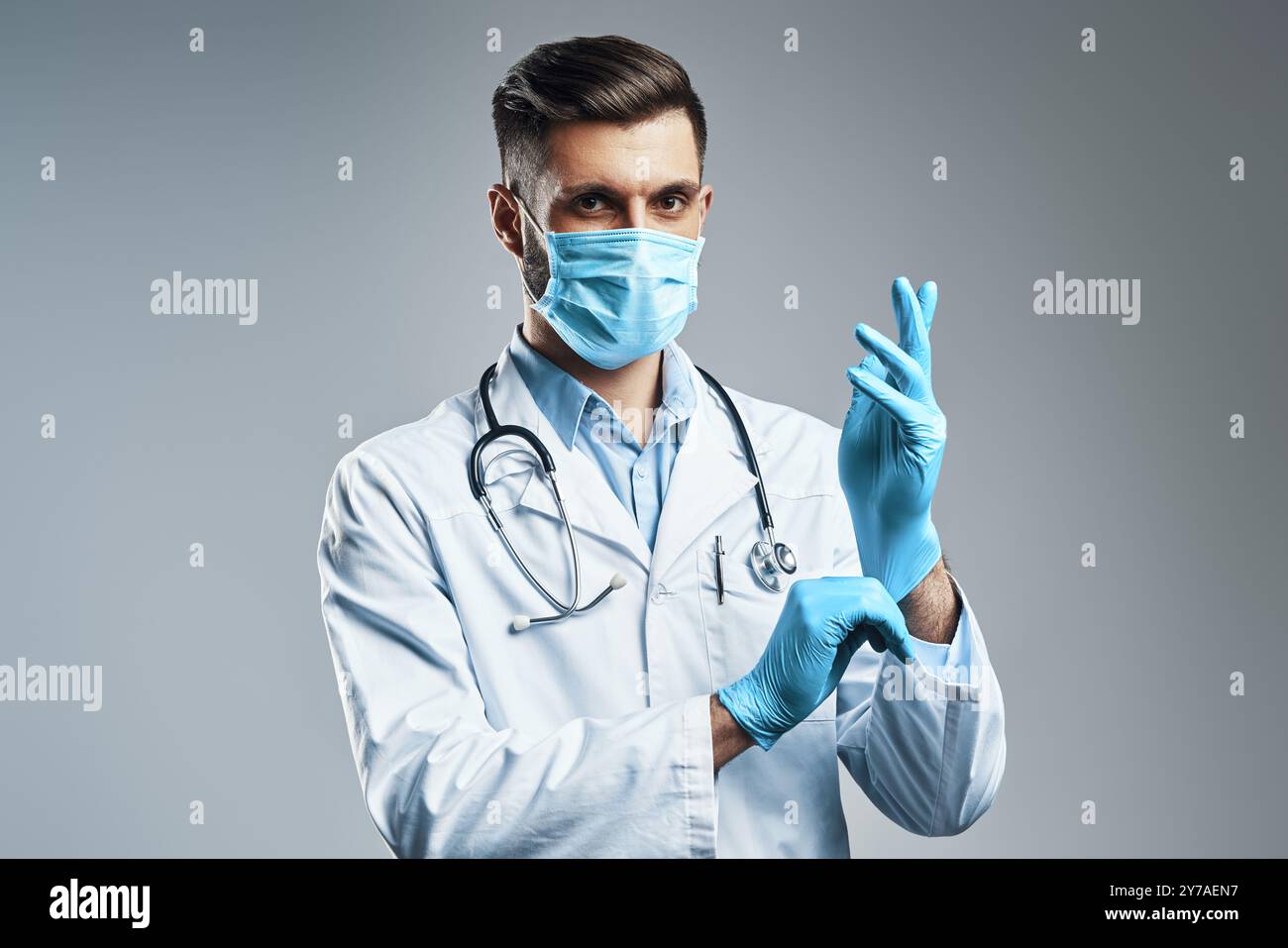 Serious young man in white lab coat and protective mask adjusting glove ...