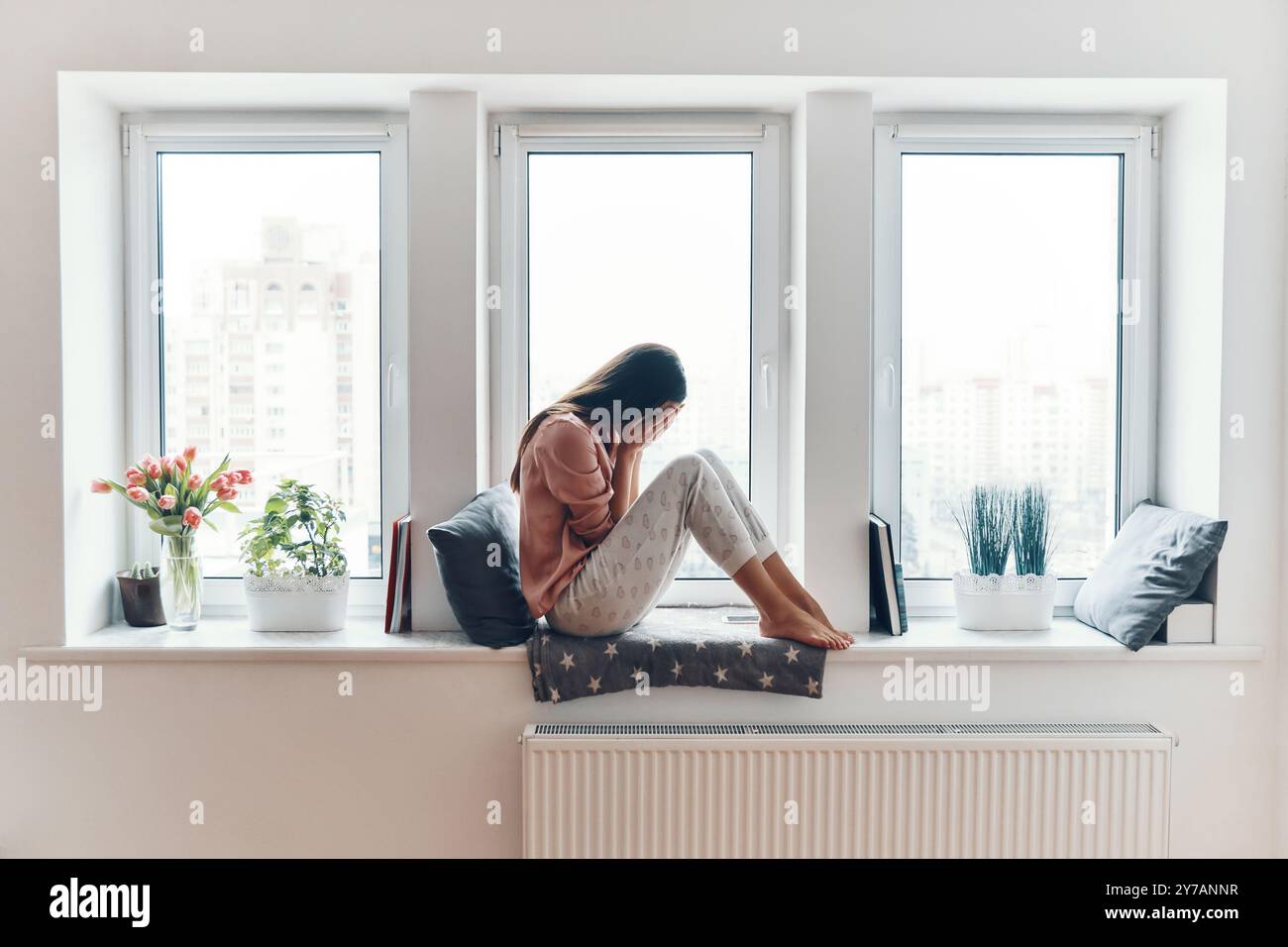 Sad young woman crying about something while sitting on the window sill ...