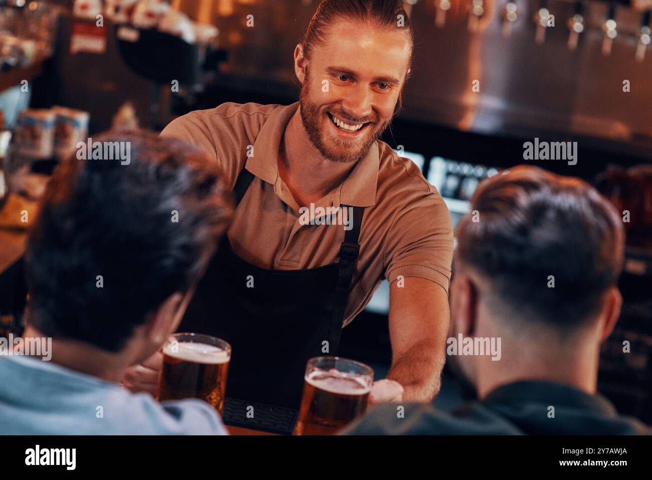Cheerful bartender serving beer to young men while standing at the bar ...