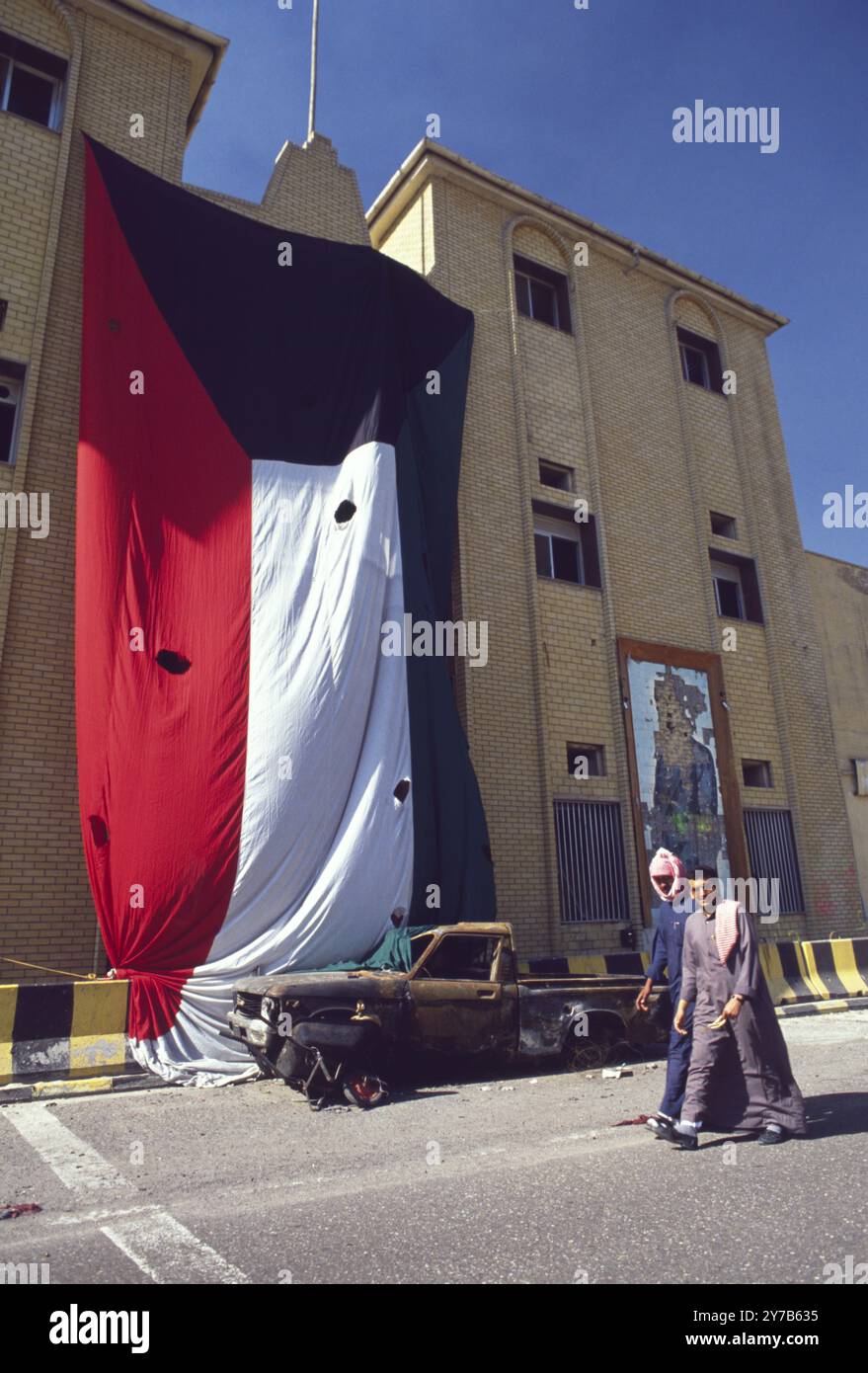8th March 1991 Two Kuwaitis walk past a huge flag of Kuwait, punctured ...