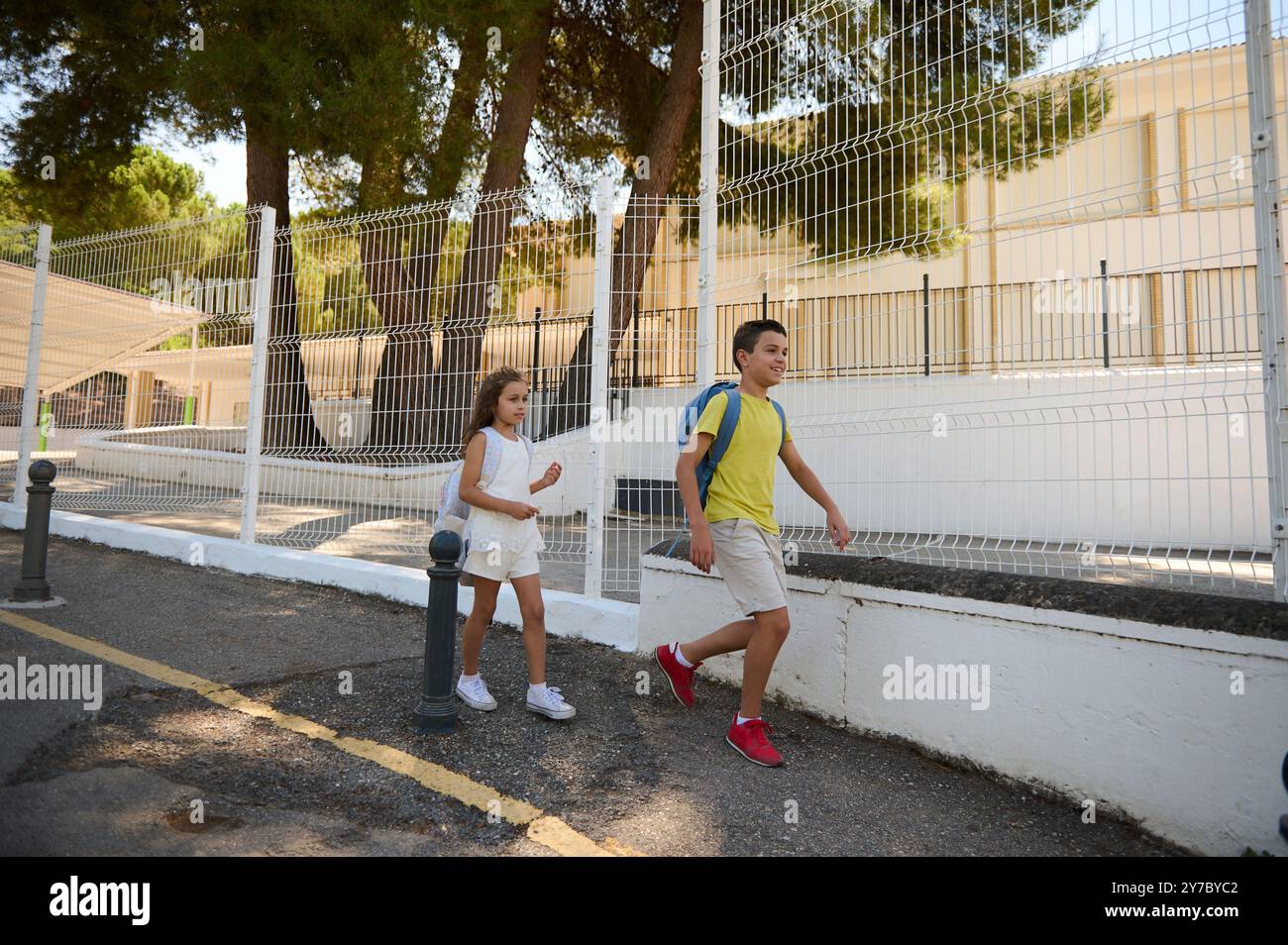 Two kids with backpacks walk joyfully along a fenced path to school ...