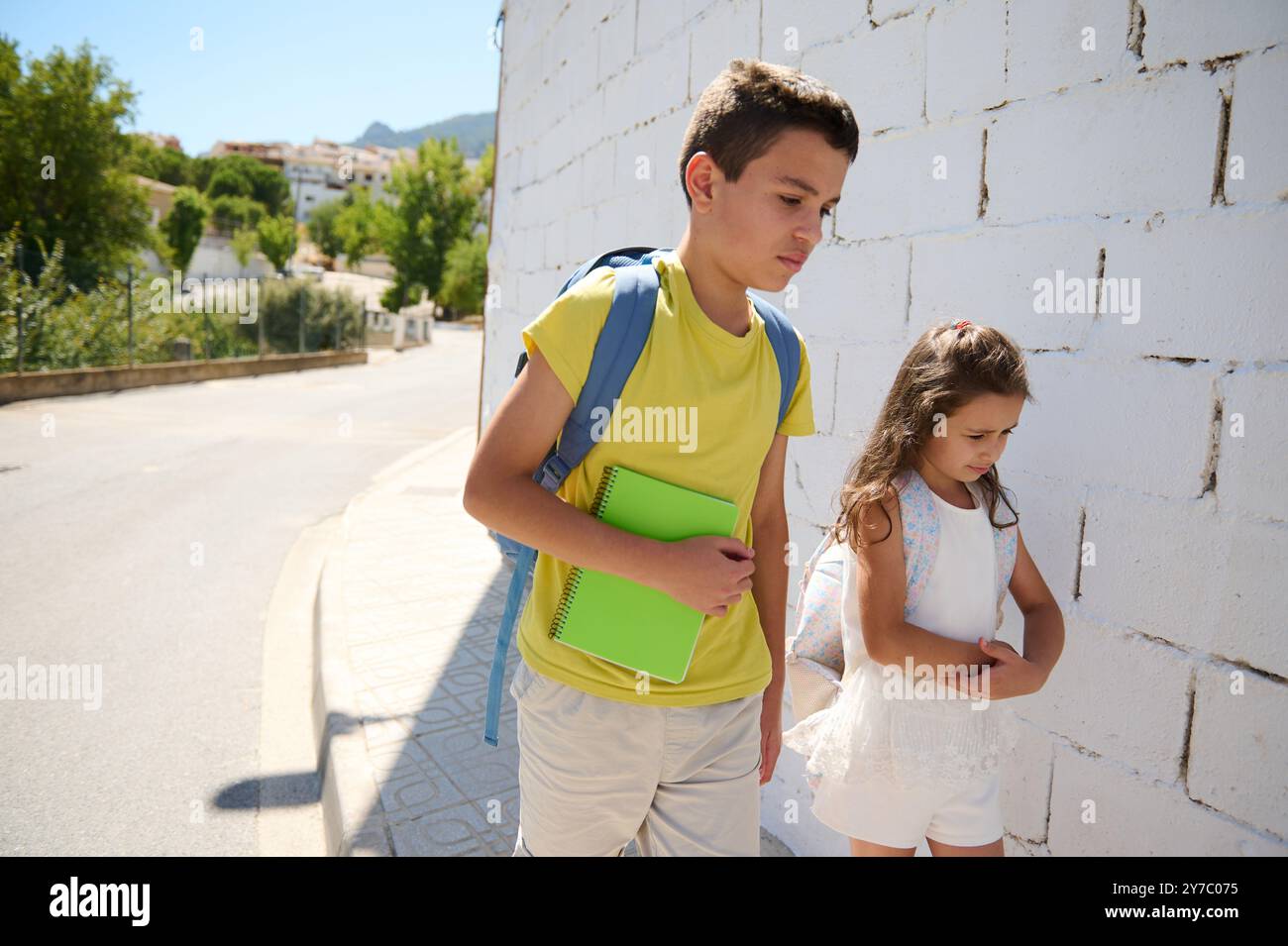 Two children with backpacks walk along a sunny street. The older ...