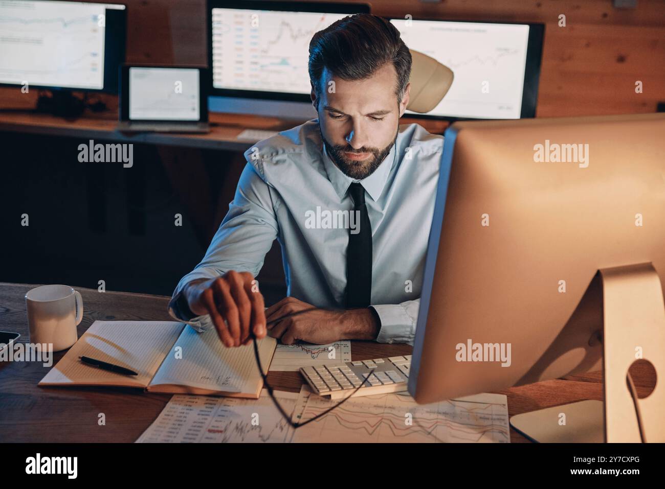 Thoughtful young man sitting at his working place while staying late in ...