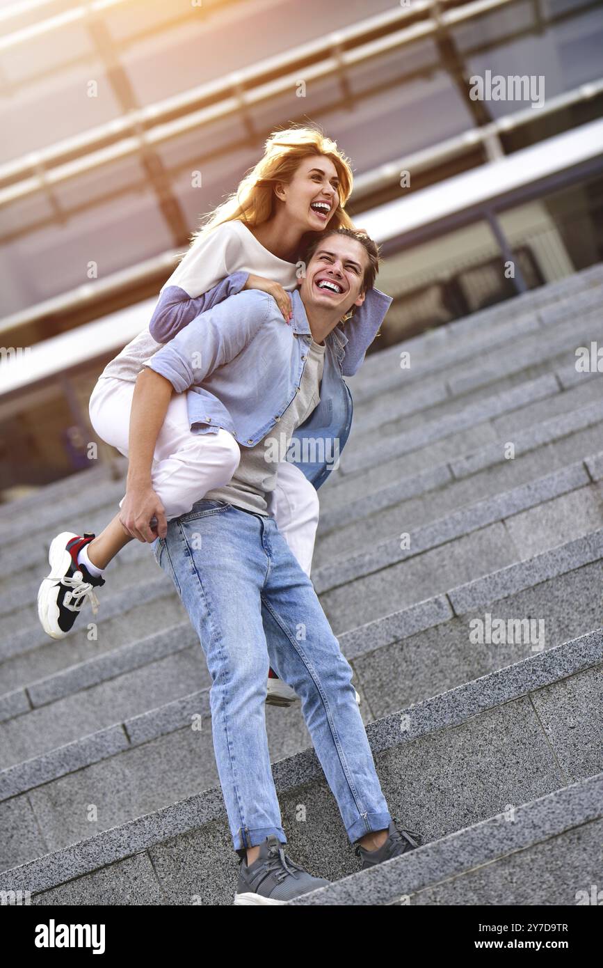 Man giving his pretty girlfriend a piggy back at street smiling at each ...