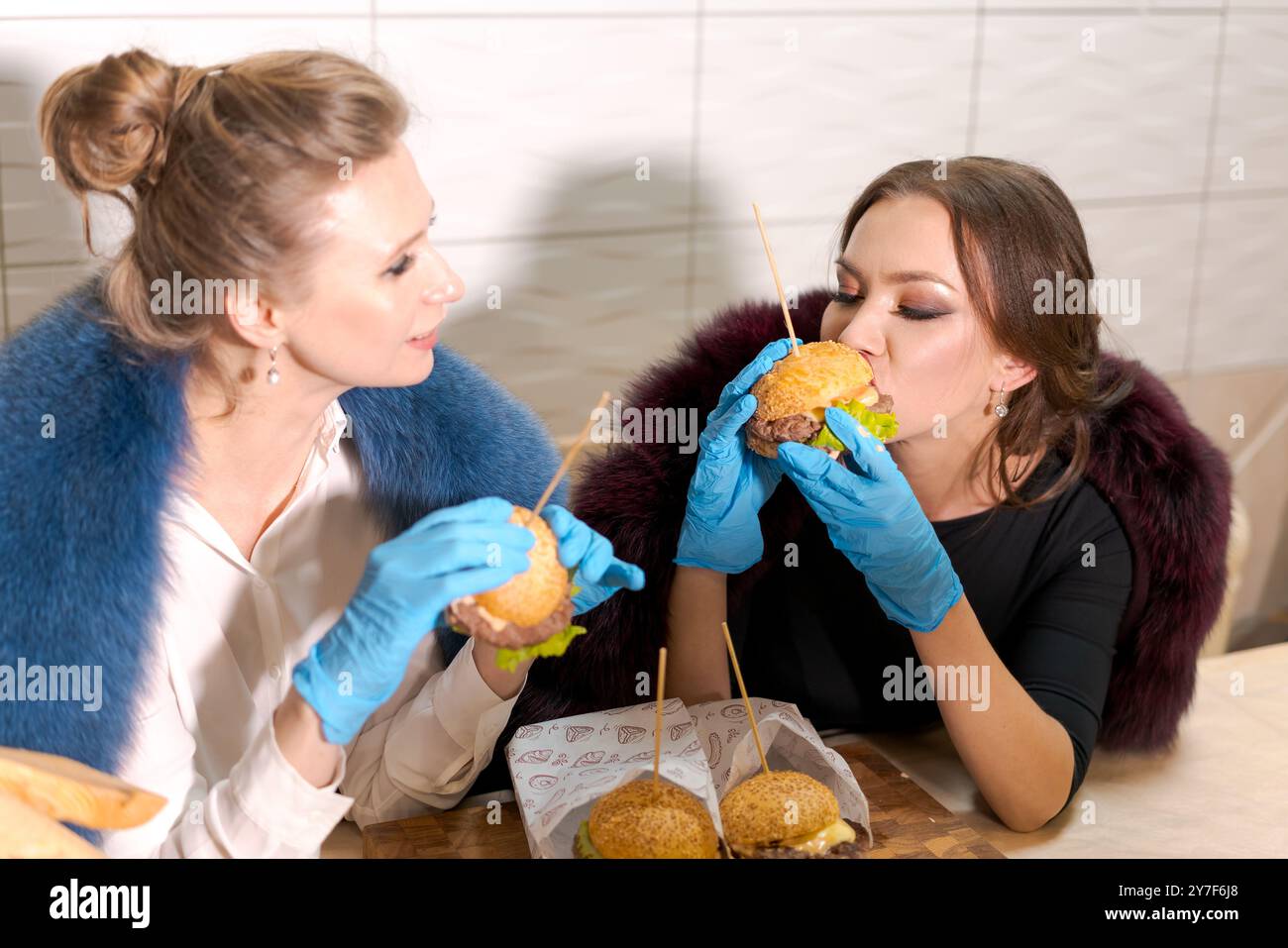 Two women eating hamburgers. One is wearing a blue fur coat. The other ...