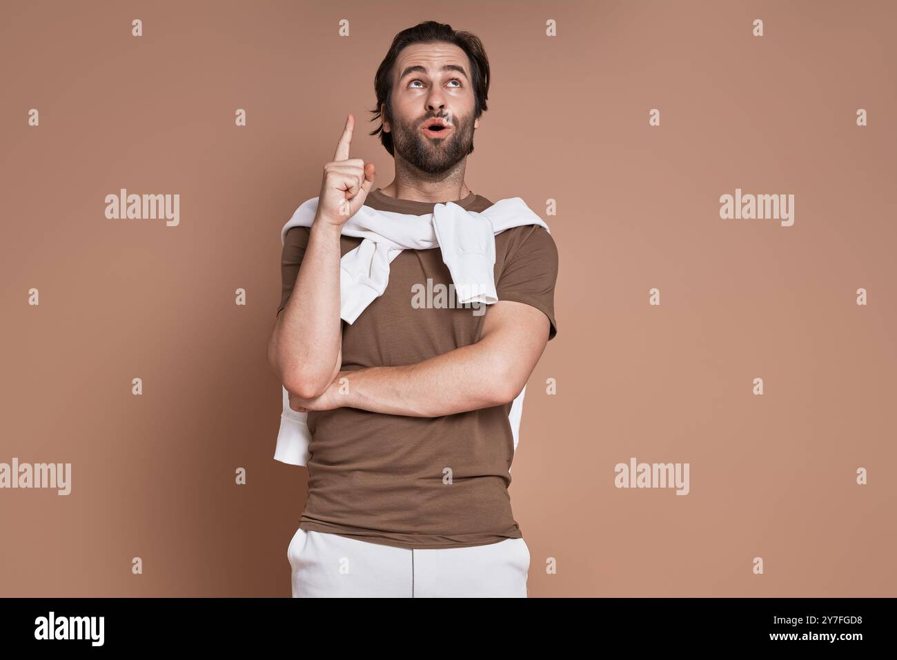 Surprised young man pointing up while standing against brown background ...