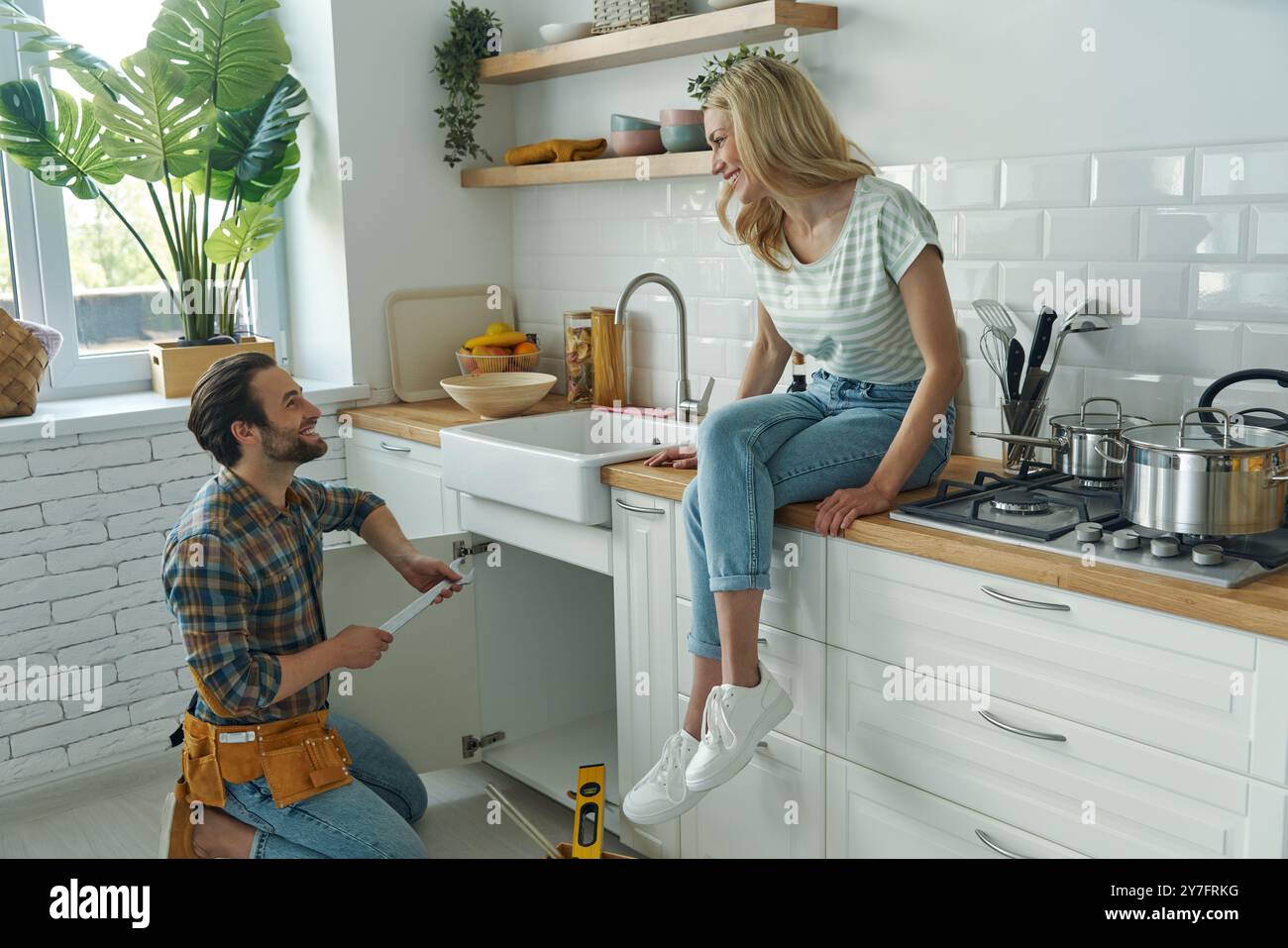 Cheerful young man repairing a sink while woman sitting on the kitchen ...