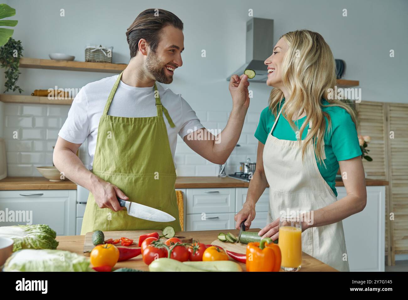 Cheerful couple feeding each other with fresh veggies while cooking at ...