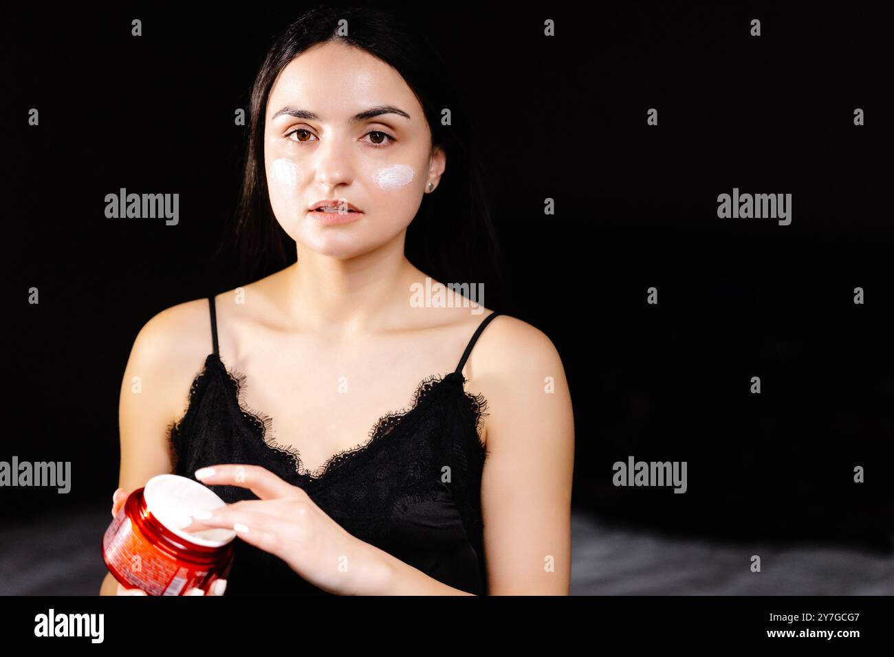 Attractive young adult woman apply facial cream look at camera ...