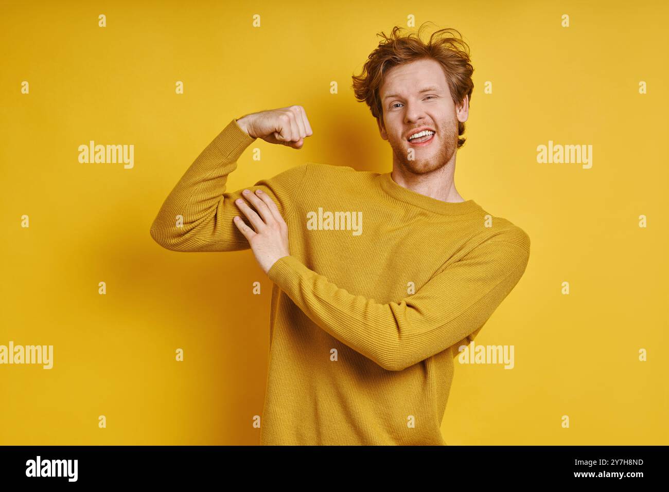 Playful redhead man touching his bicep while standing against yellow ...