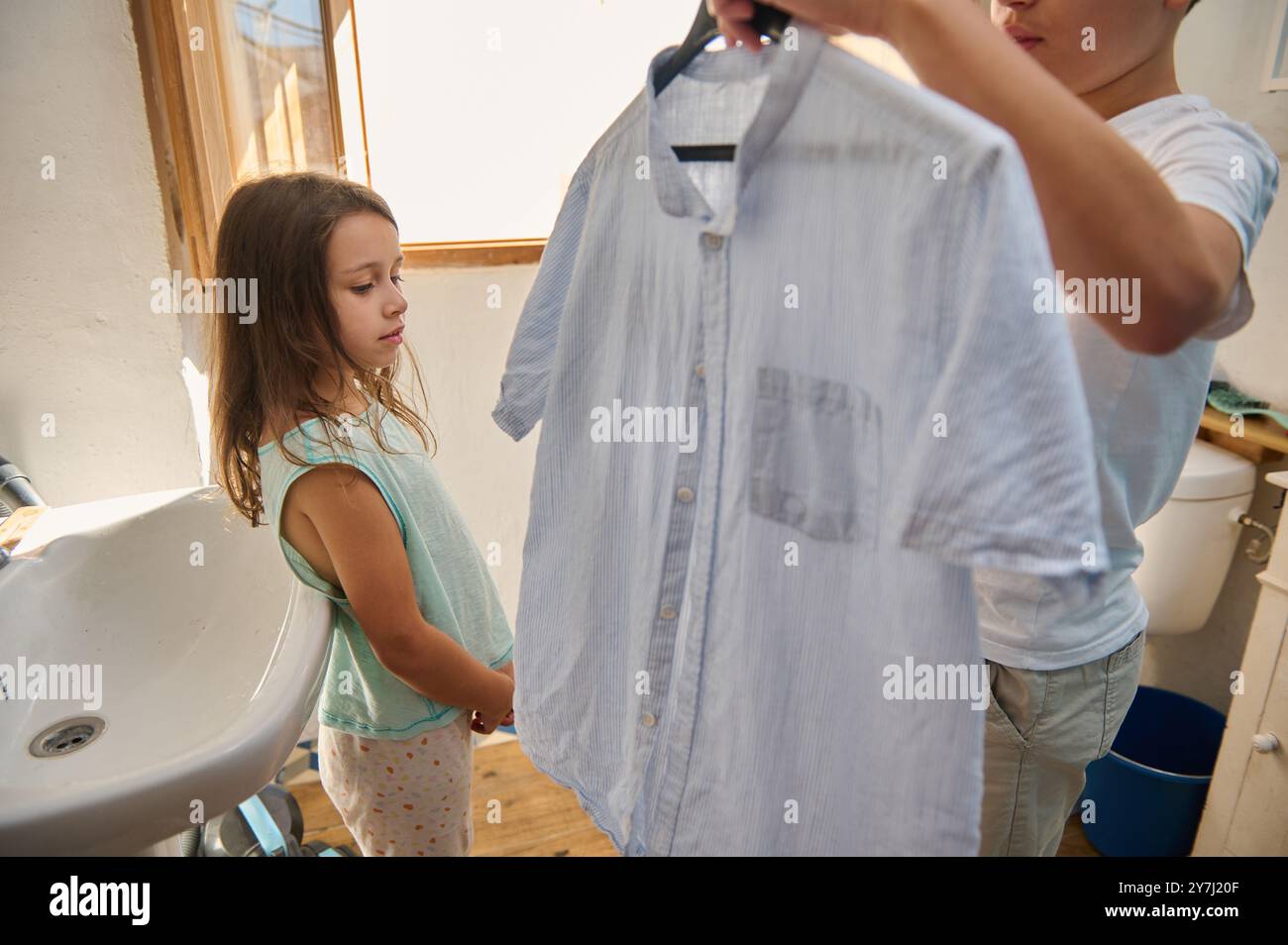 A young girl and boy engage in laundry tasks in a sunlit bathroom ...