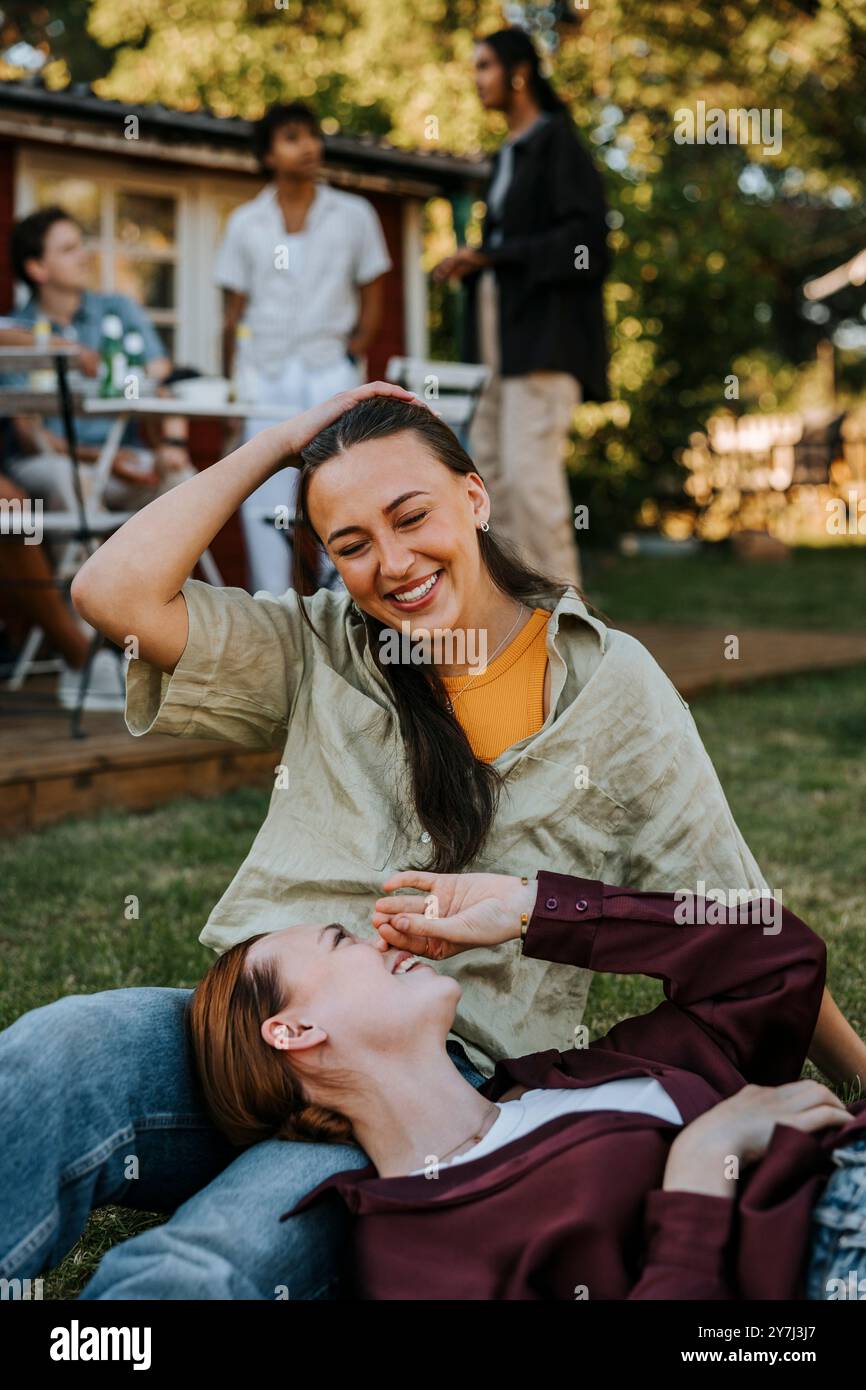 Happy woman lying down on lap of female friend sitting on grass in back ...