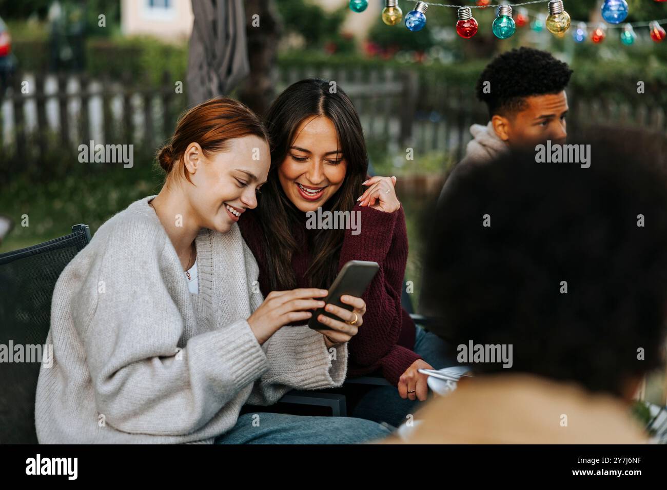 Female friends using smart phone while sitting on chair in back yard at ...