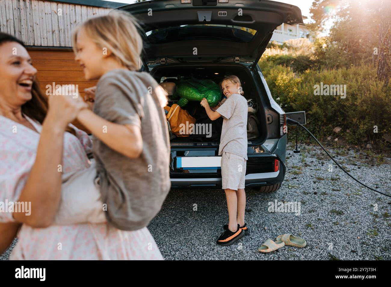 Boy loading car trunk and looking at mother playing with sibling on ...