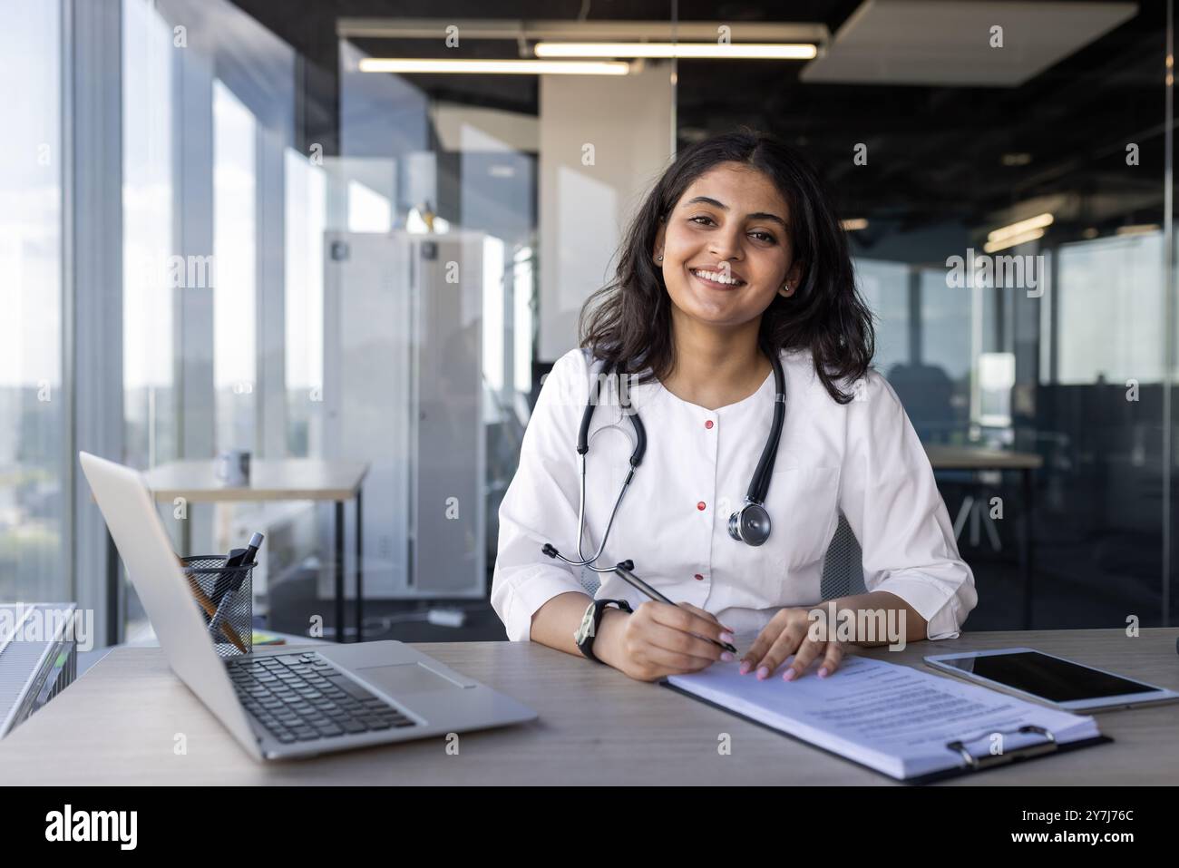 Smiling female doctor with stethoscope at desk working with laptop ...