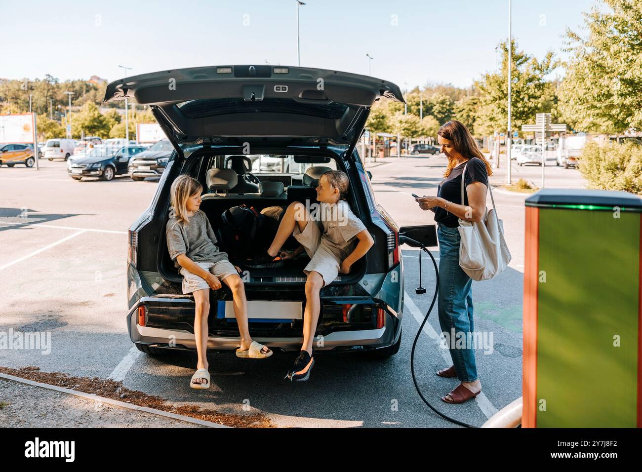 Mother using smart phone with sons sitting in trunk while charging car ...