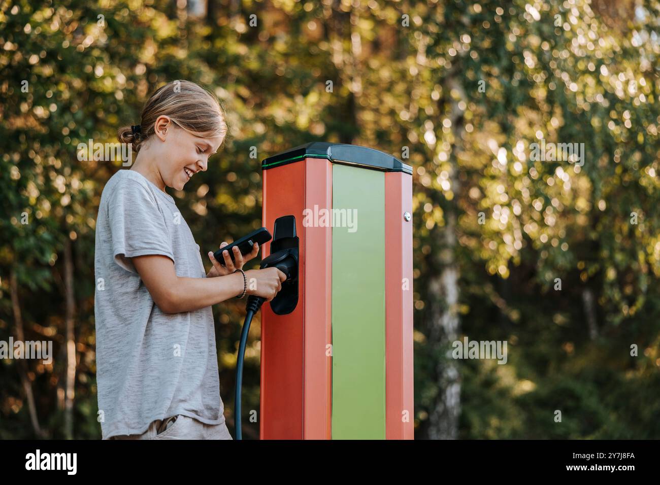 Happy boy using smart phone while holding electric plug at car charging ...