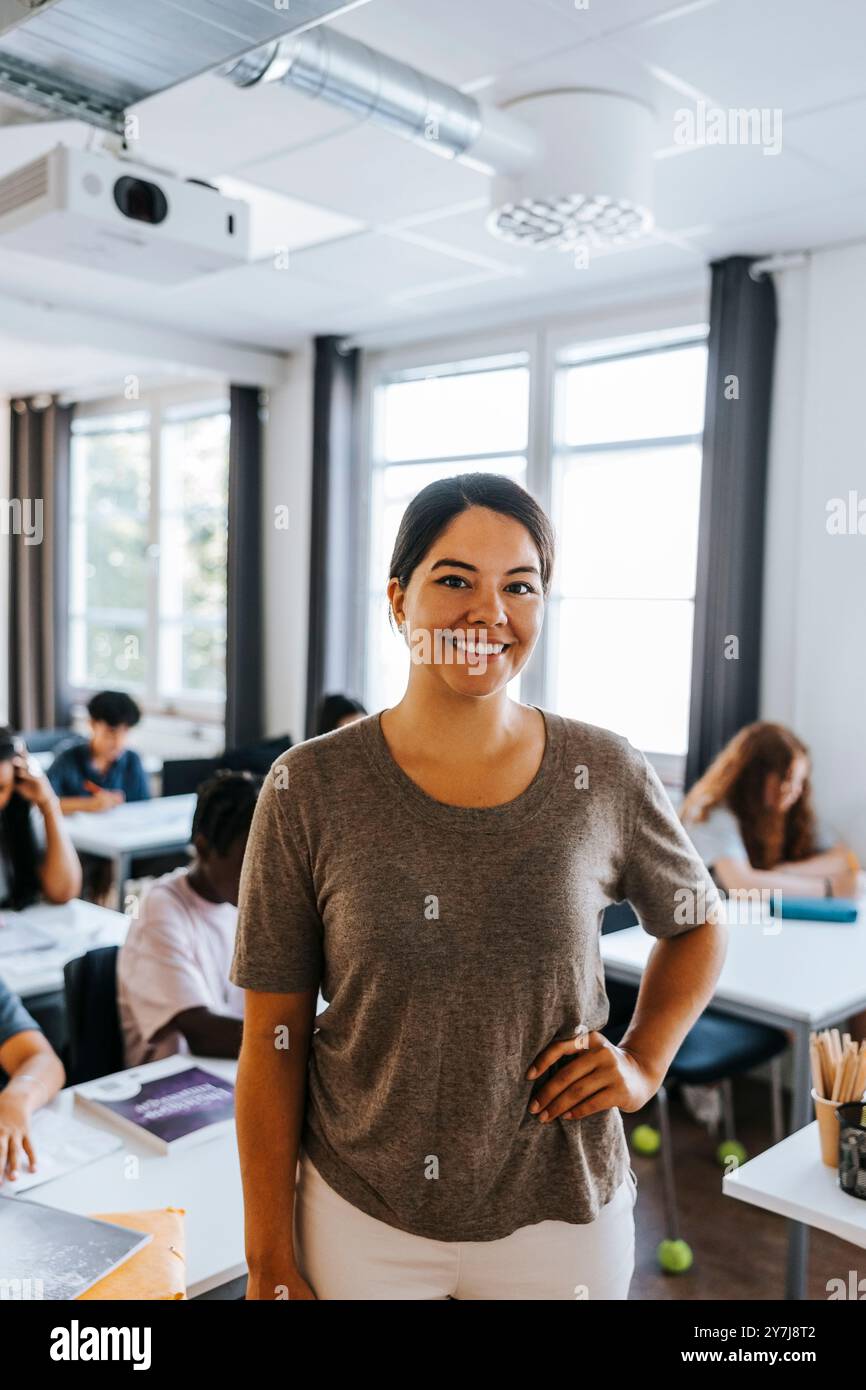 Portrait of happy female teacher with hand on hip standing in classroom ...