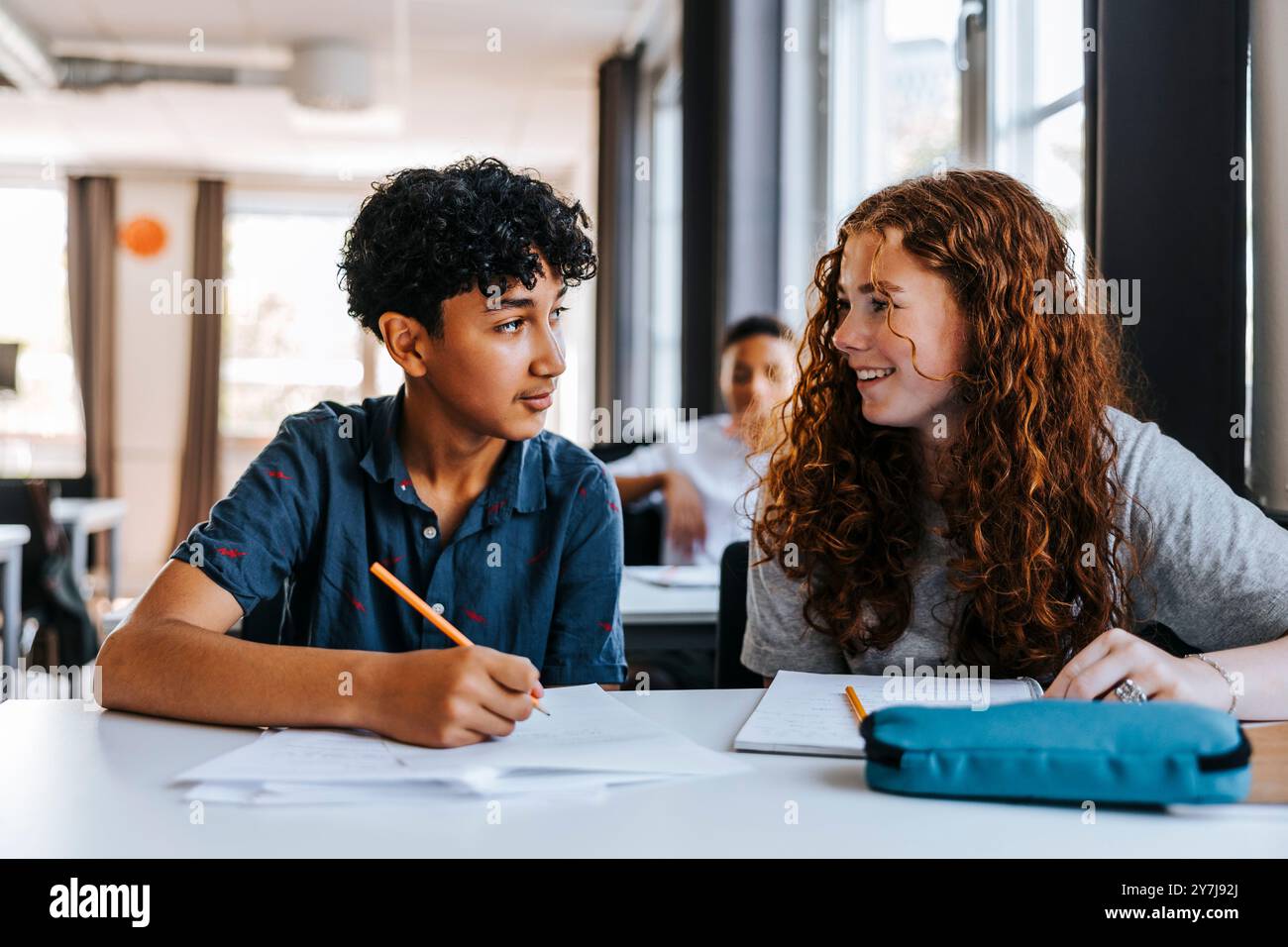 Smiling curly hair teenage girl studying with male classmate while ...