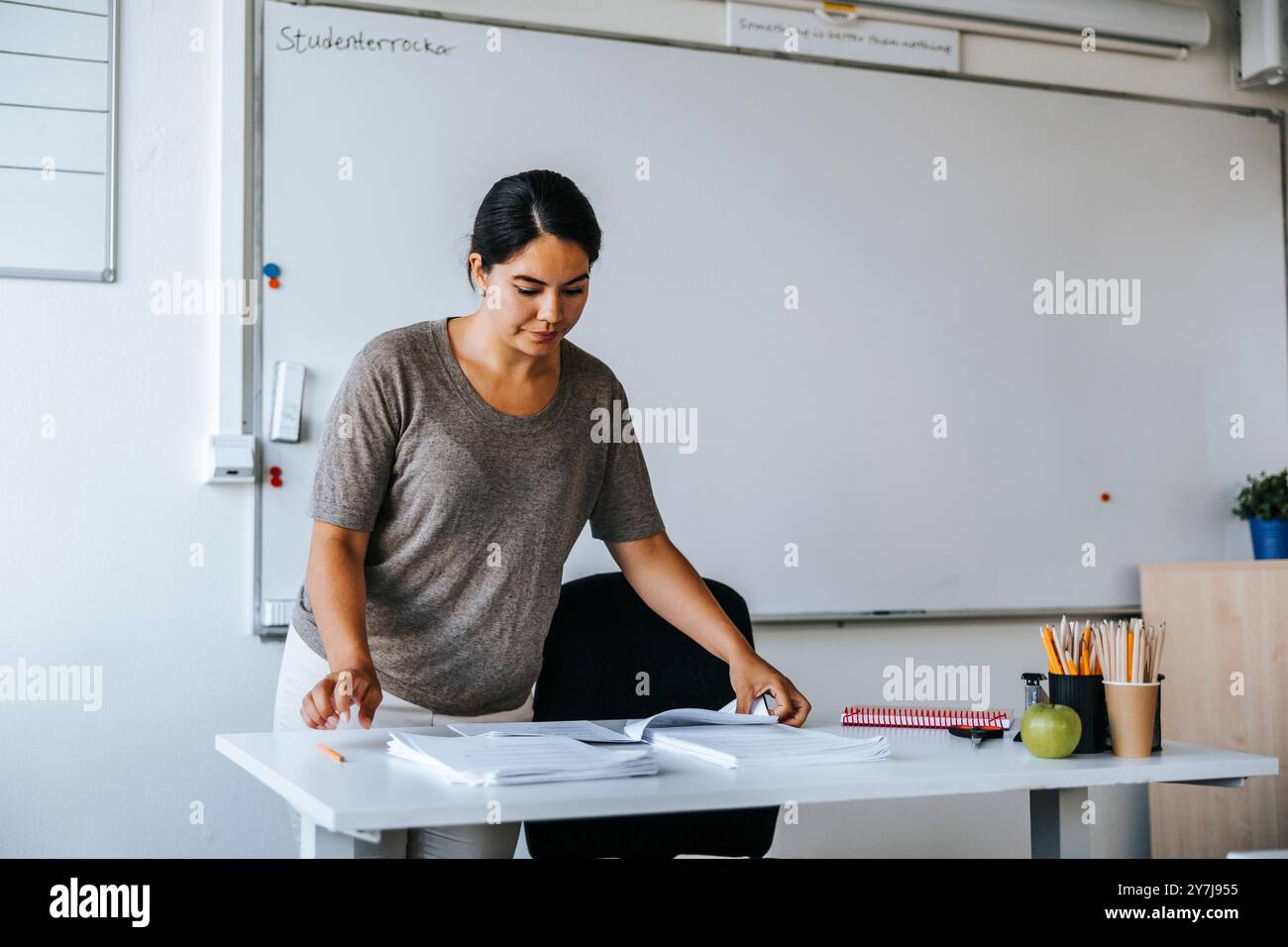 Focused female teacher preparing test papers while standing near table ...