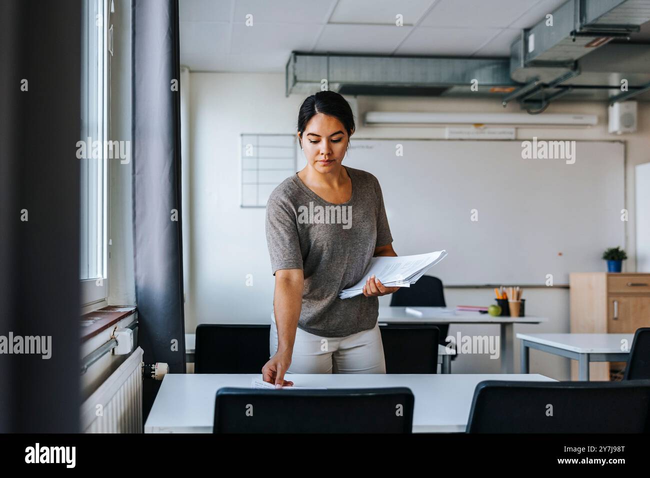 Female teacher keeping test papers on desk while standing in classroom ...