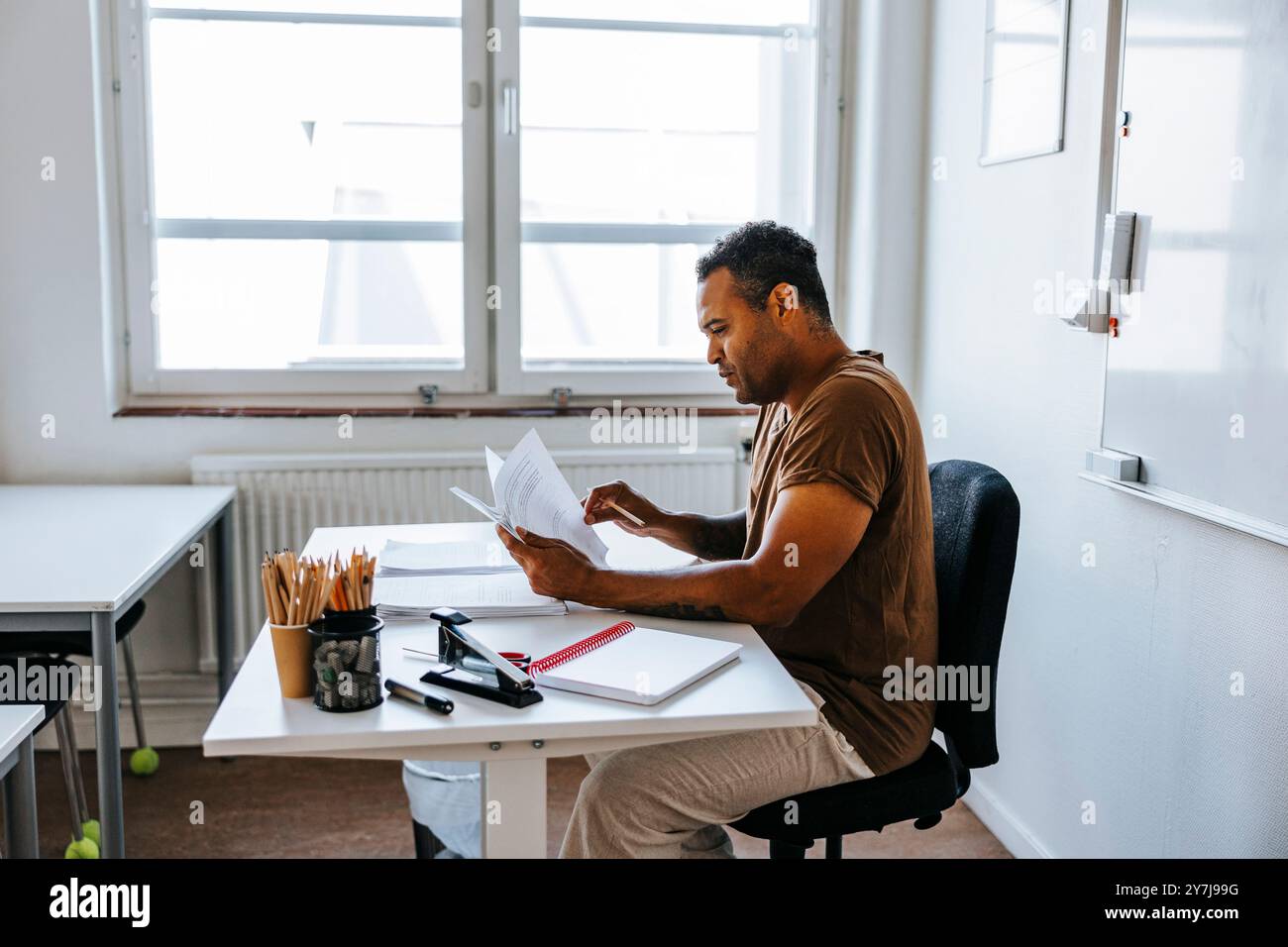 Focused male teacher checking test papers while sitting near table in ...