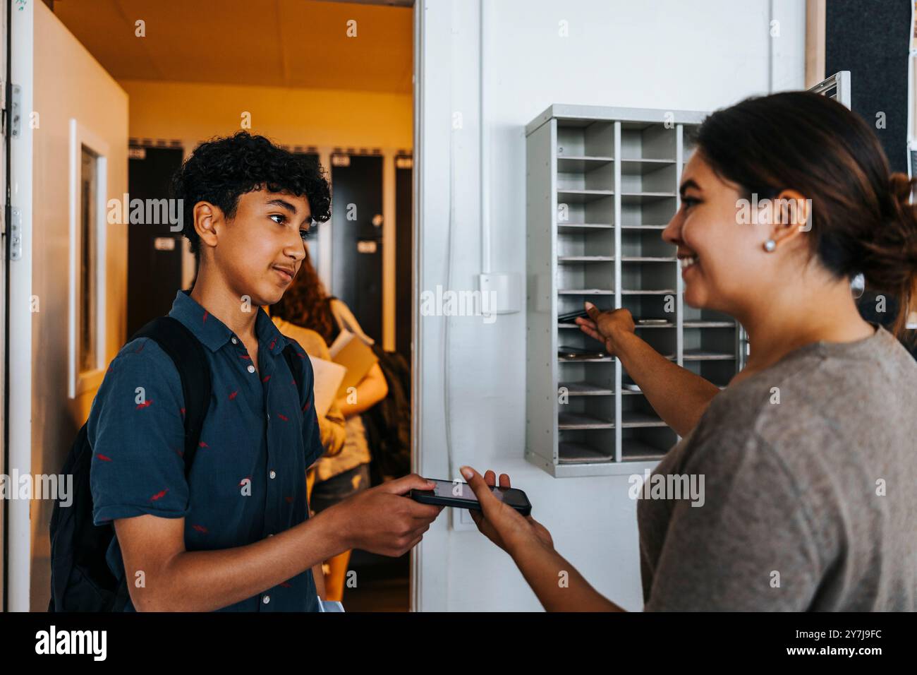 Smiling female teacher collecting smart phone from teenage boy standing ...