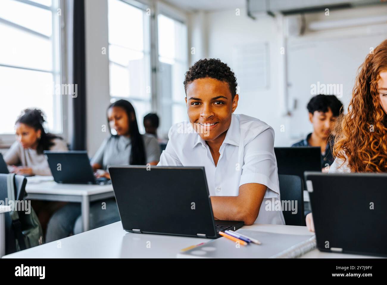 Portrait of smiling teenage boy sitting with laptop near desk in ...
