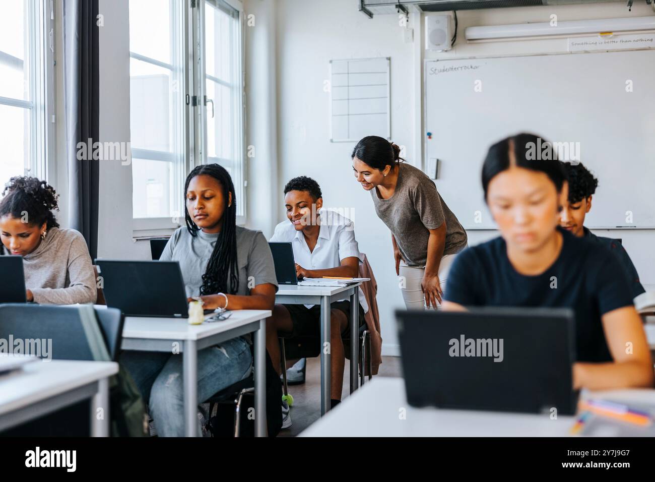 Smiling female teacher assisting student studying on laptop while ...
