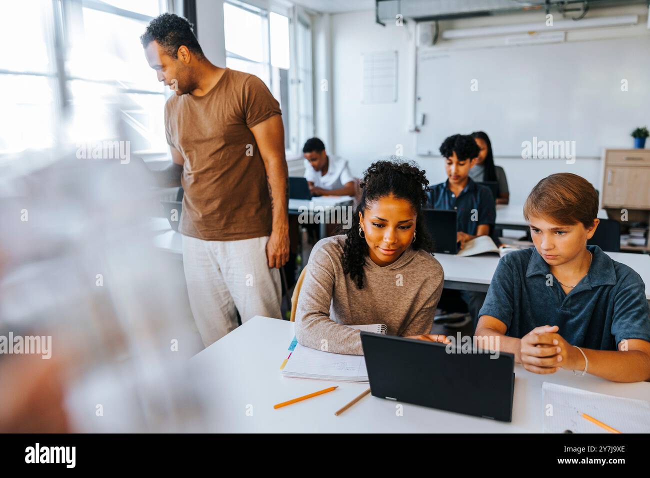 Teacher standing near male and female students doing e-learning in ...