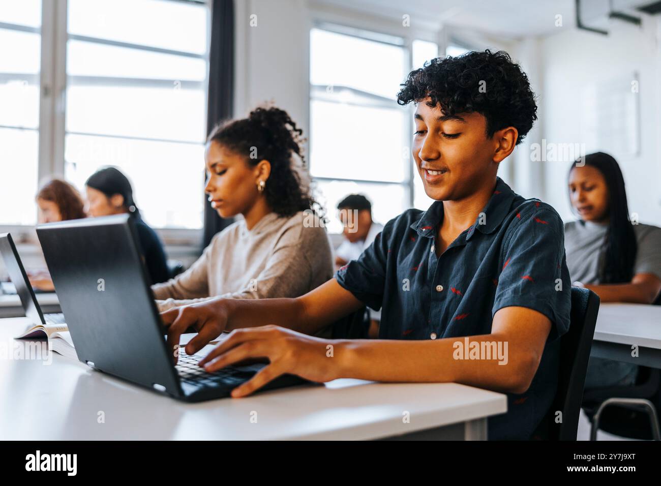 Smiling teenage boy using laptop while sitting with students in ...
