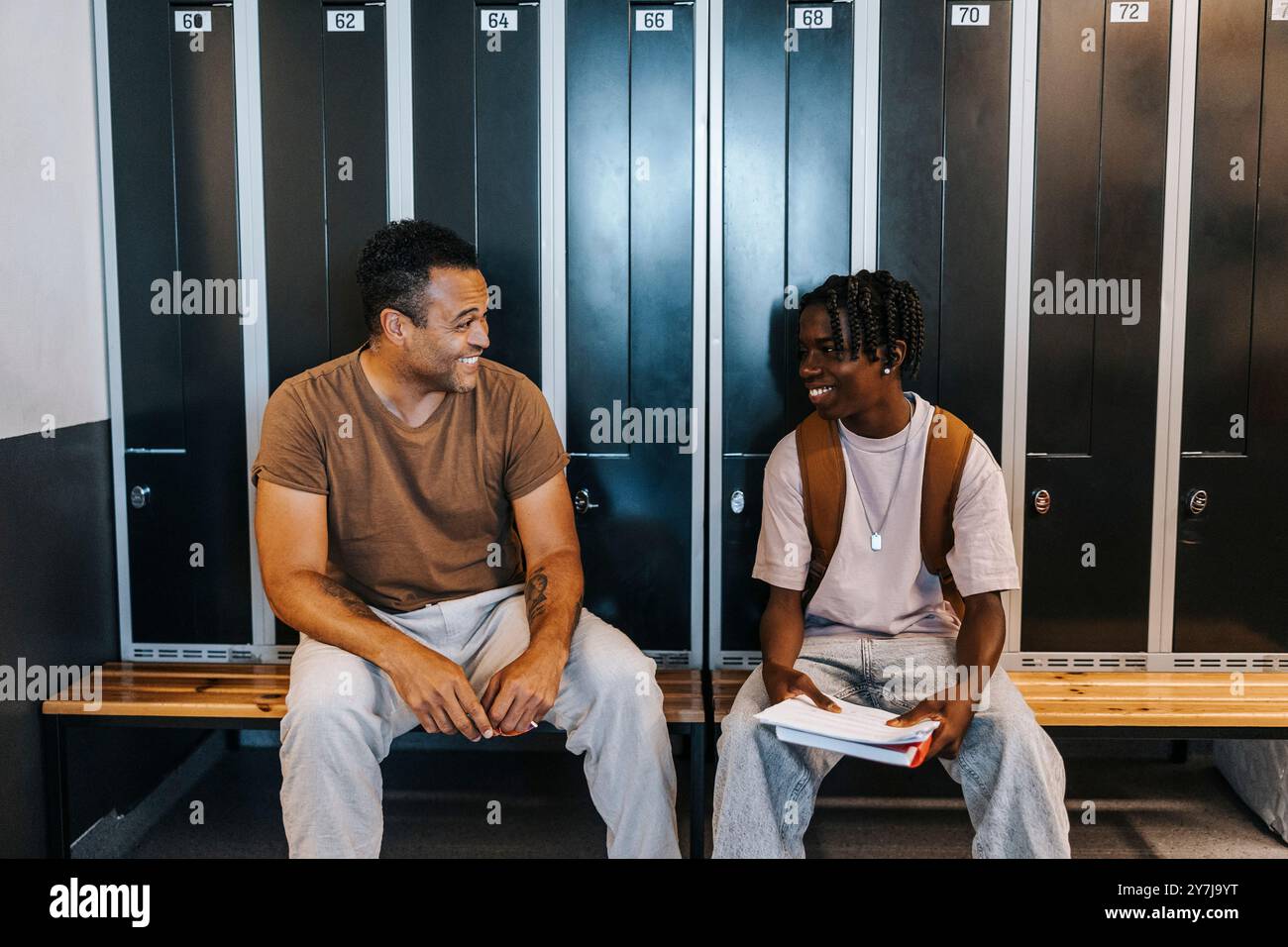 Happy male couch and teenage boy sitting on bench in locker room at ...