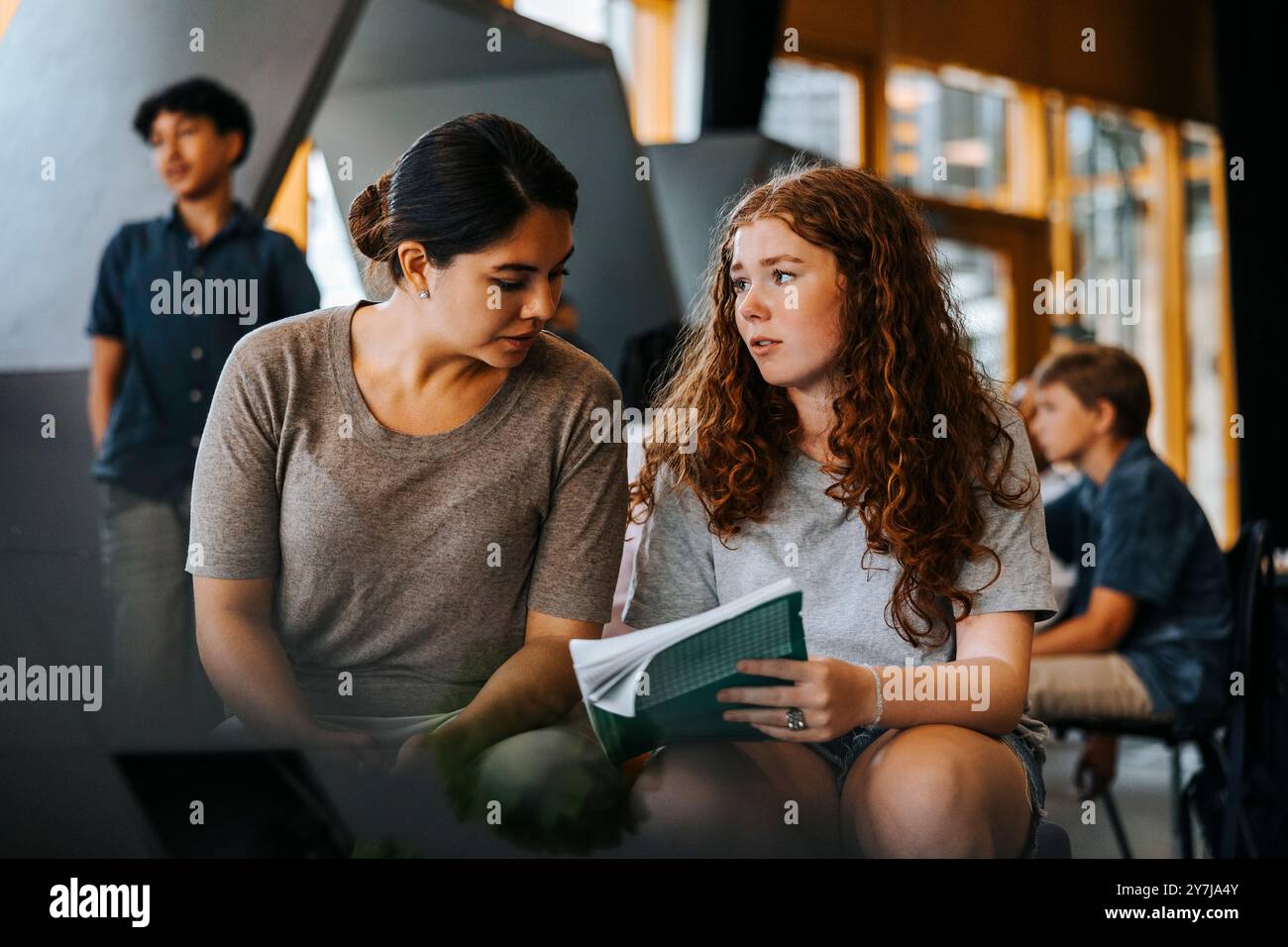Female teacher assisting teenage girl holding book while sitting in ...