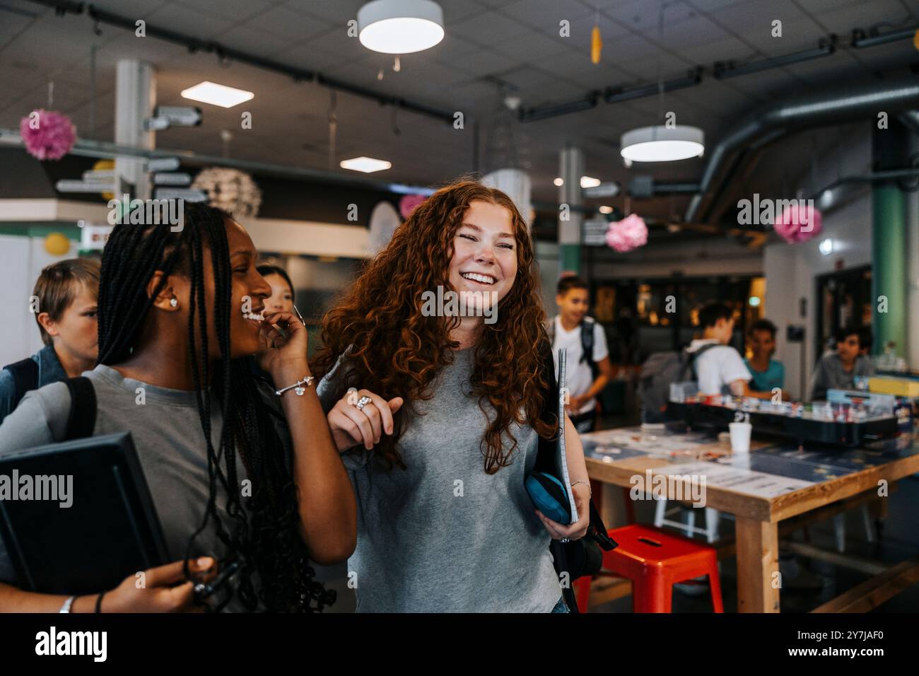 Happy curly haired teenage girl walking with female friend in cafeteria ...