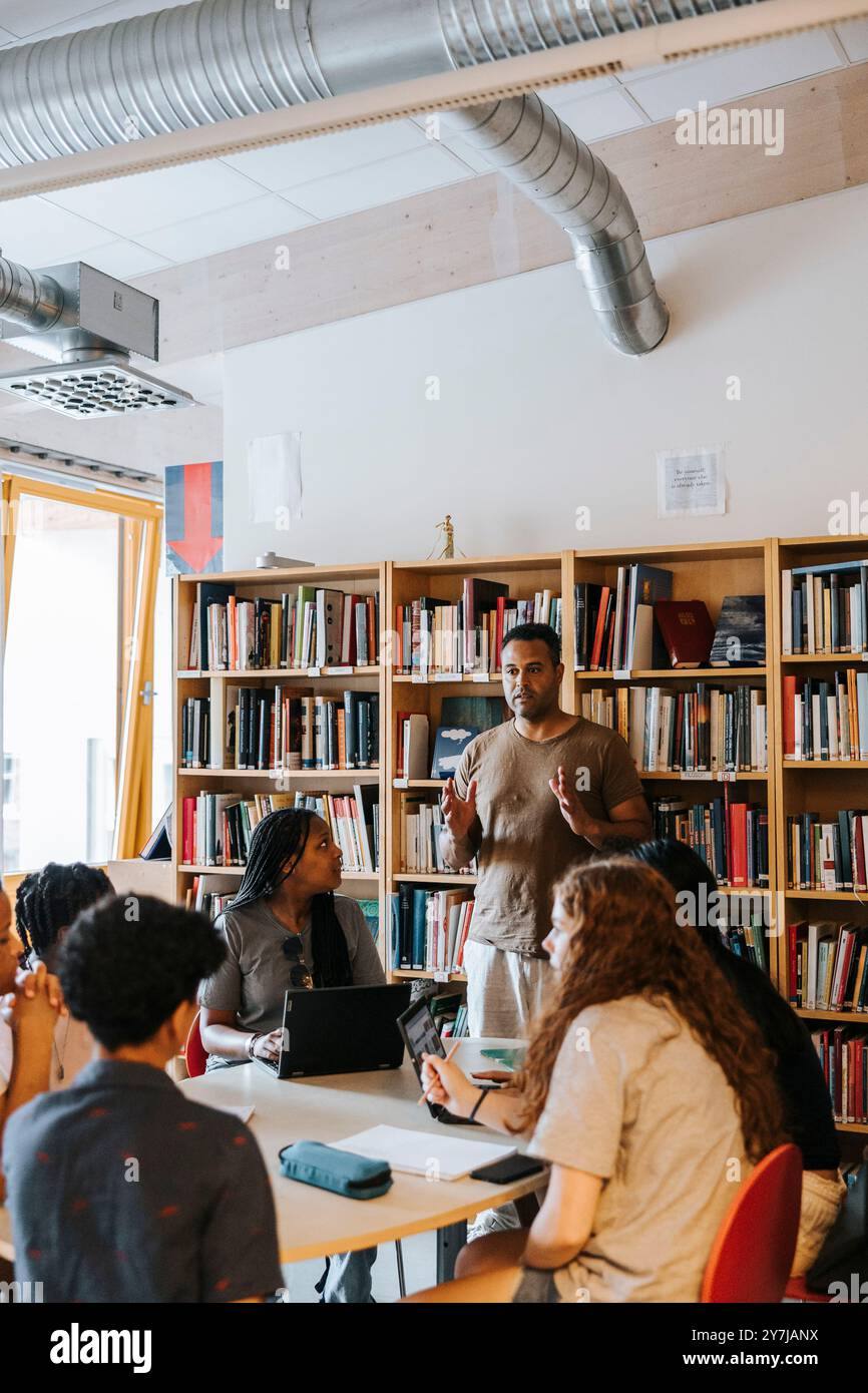 Male librarian explaining junior high students sitting near table in ...