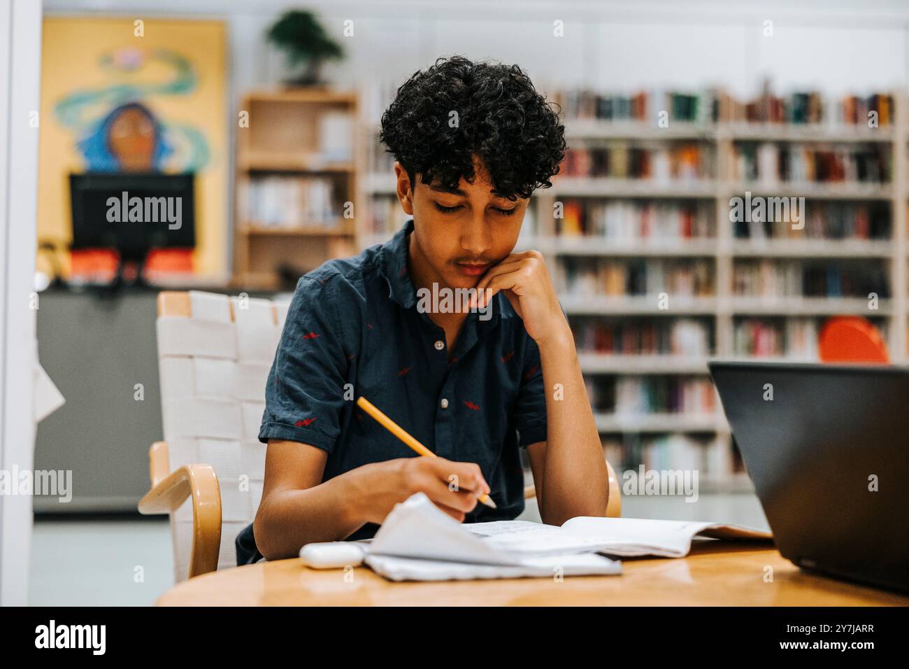 Focused teenage boy studying with hand on chin while sitting near table ...