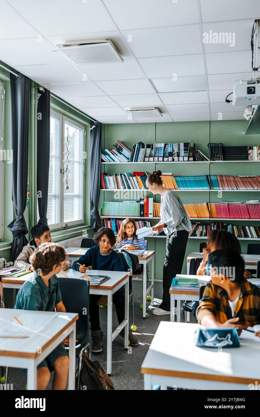 Side view of female professor giving test papers to elementary students ...