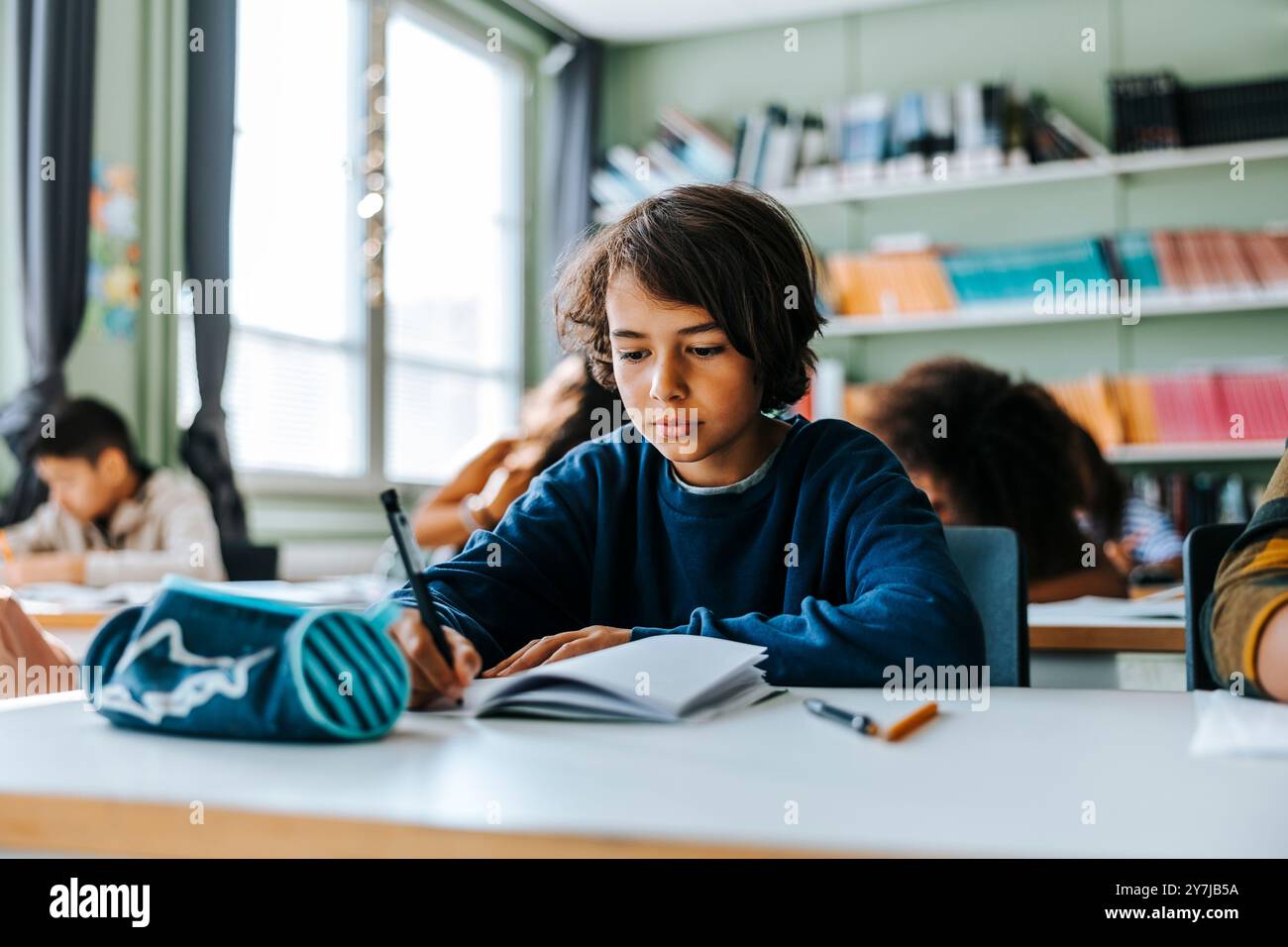Focused boy writing in book while studying in classroom at elementary ...