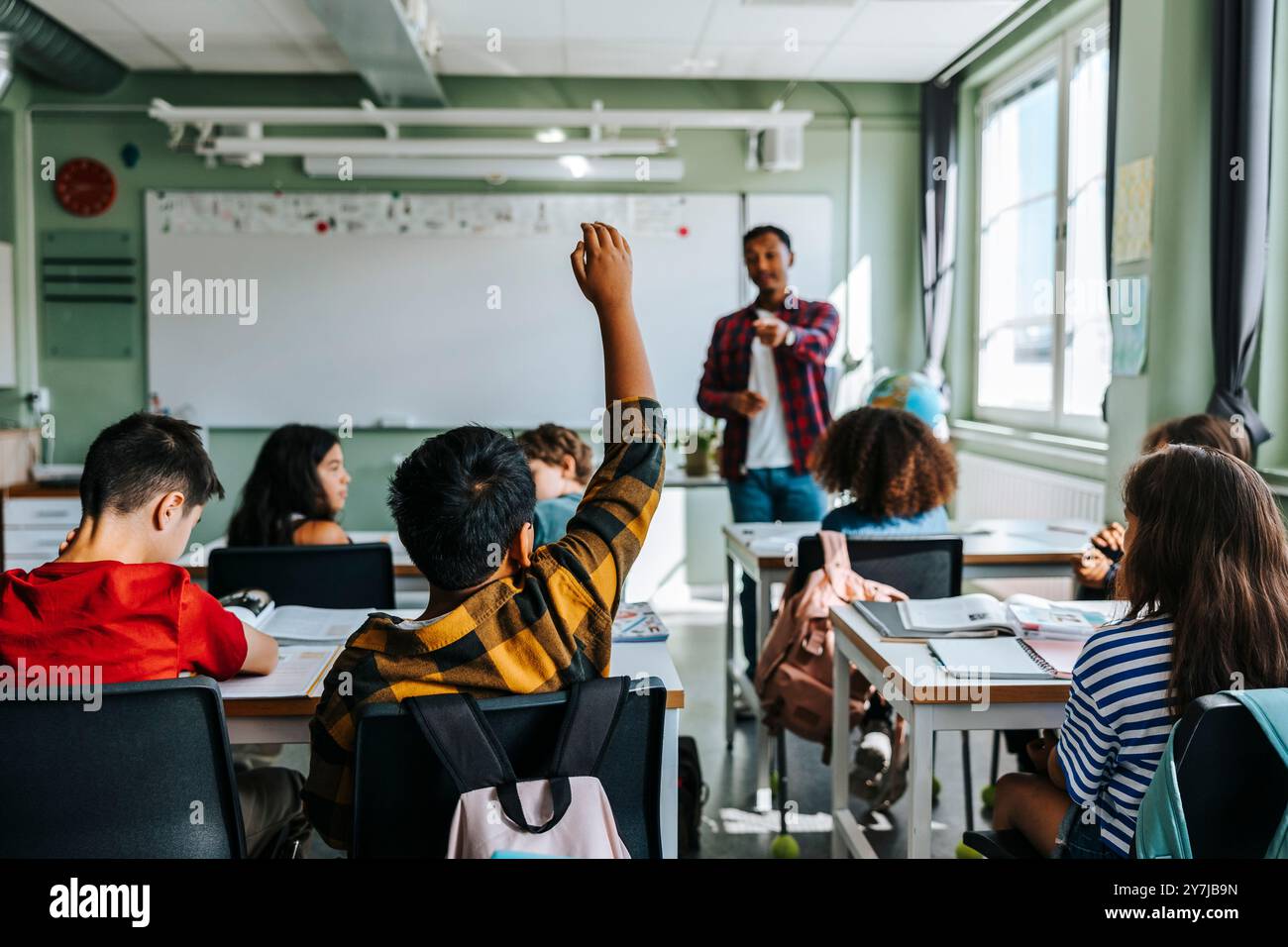 Rear view of boy raising hand for asking question to male teacher while ...