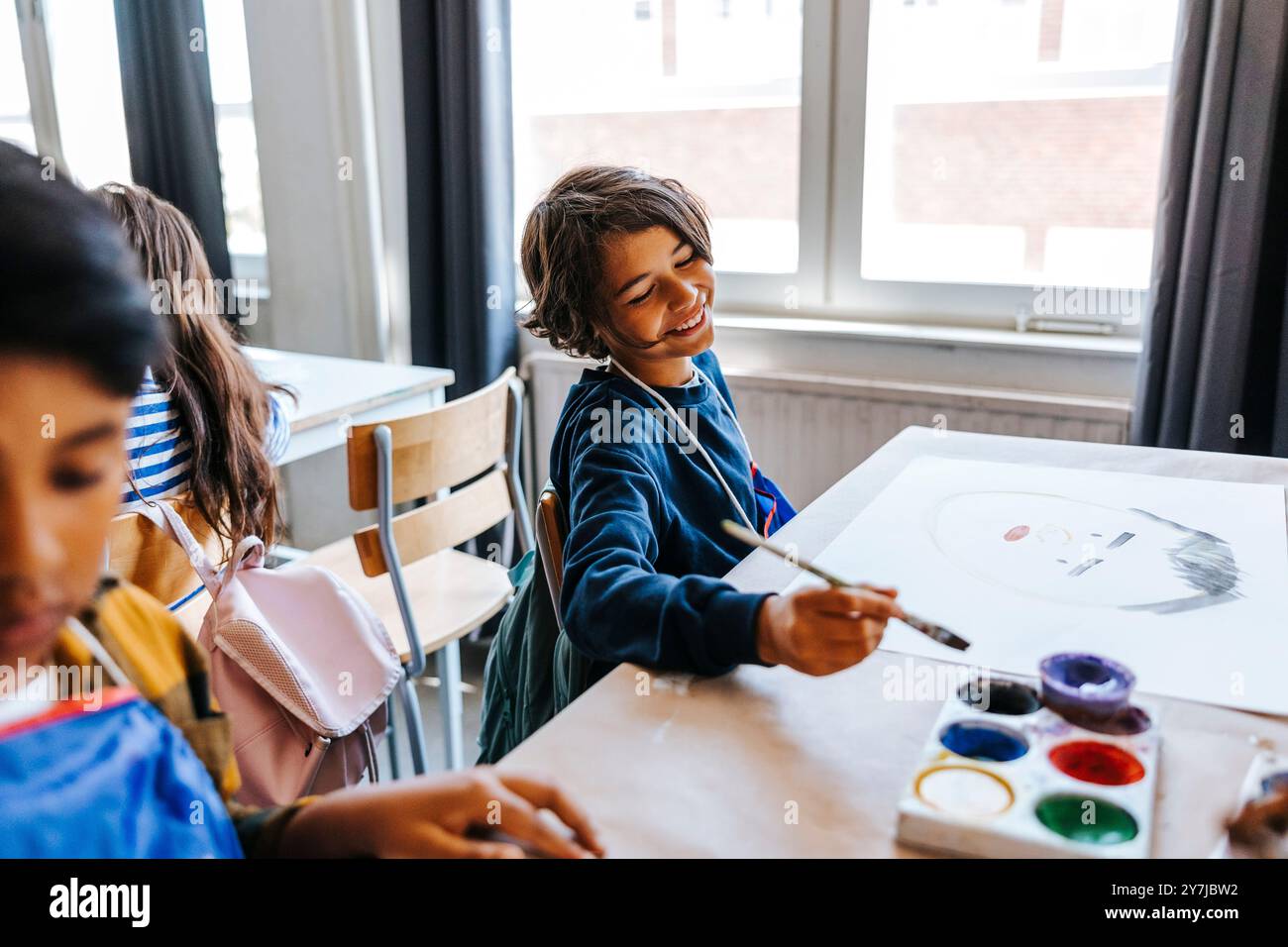 Happy boy painting with paintbrush while sitting near desk in art class ...
