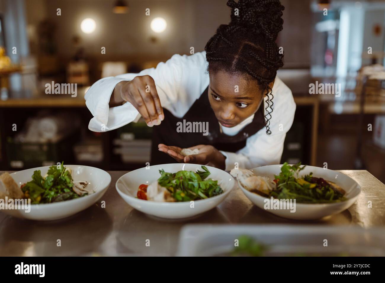 Focused female chef sprinkling salt on salad while bending in ...