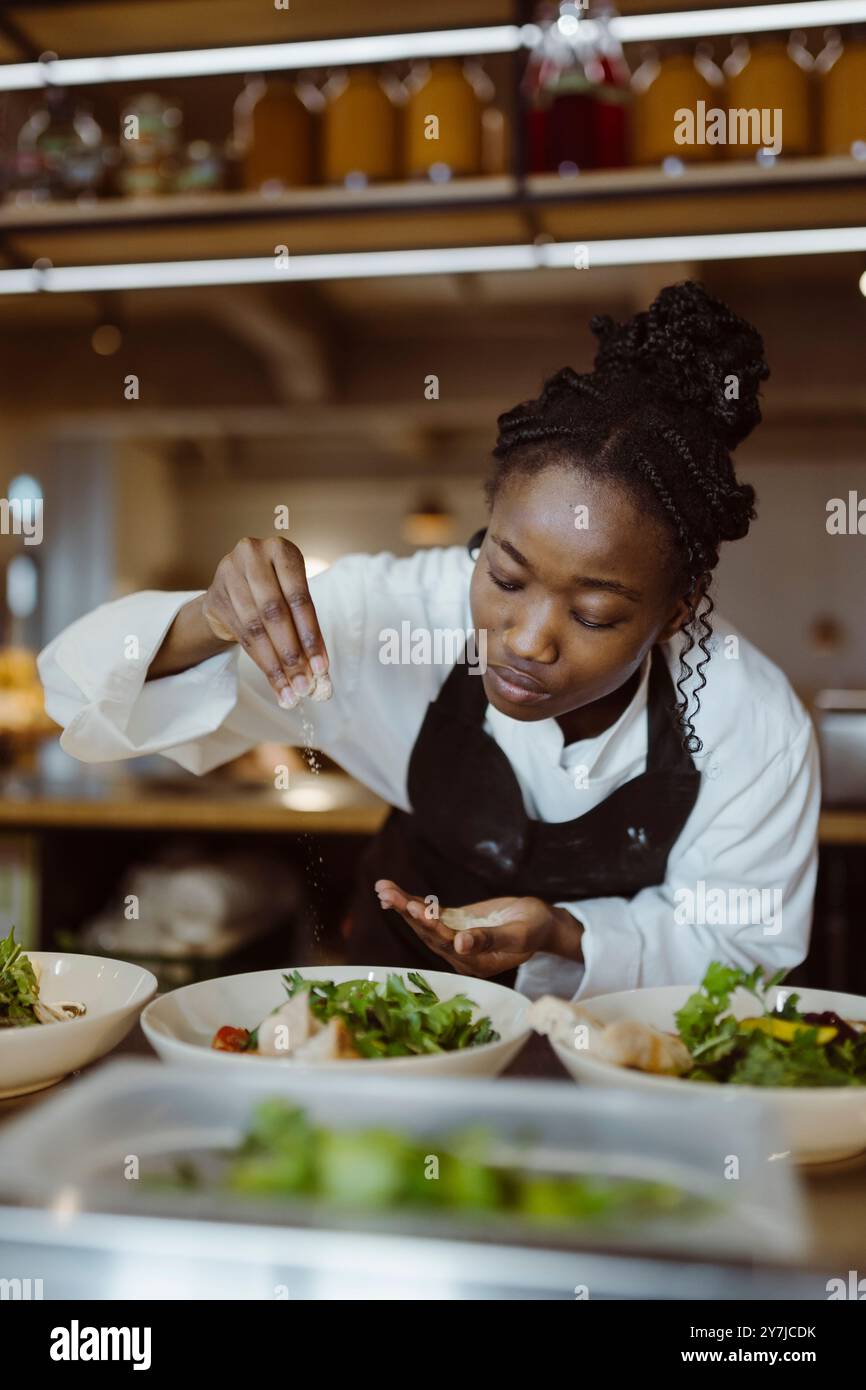 Focused young female chef sprinkling salt on salad while bending in ...