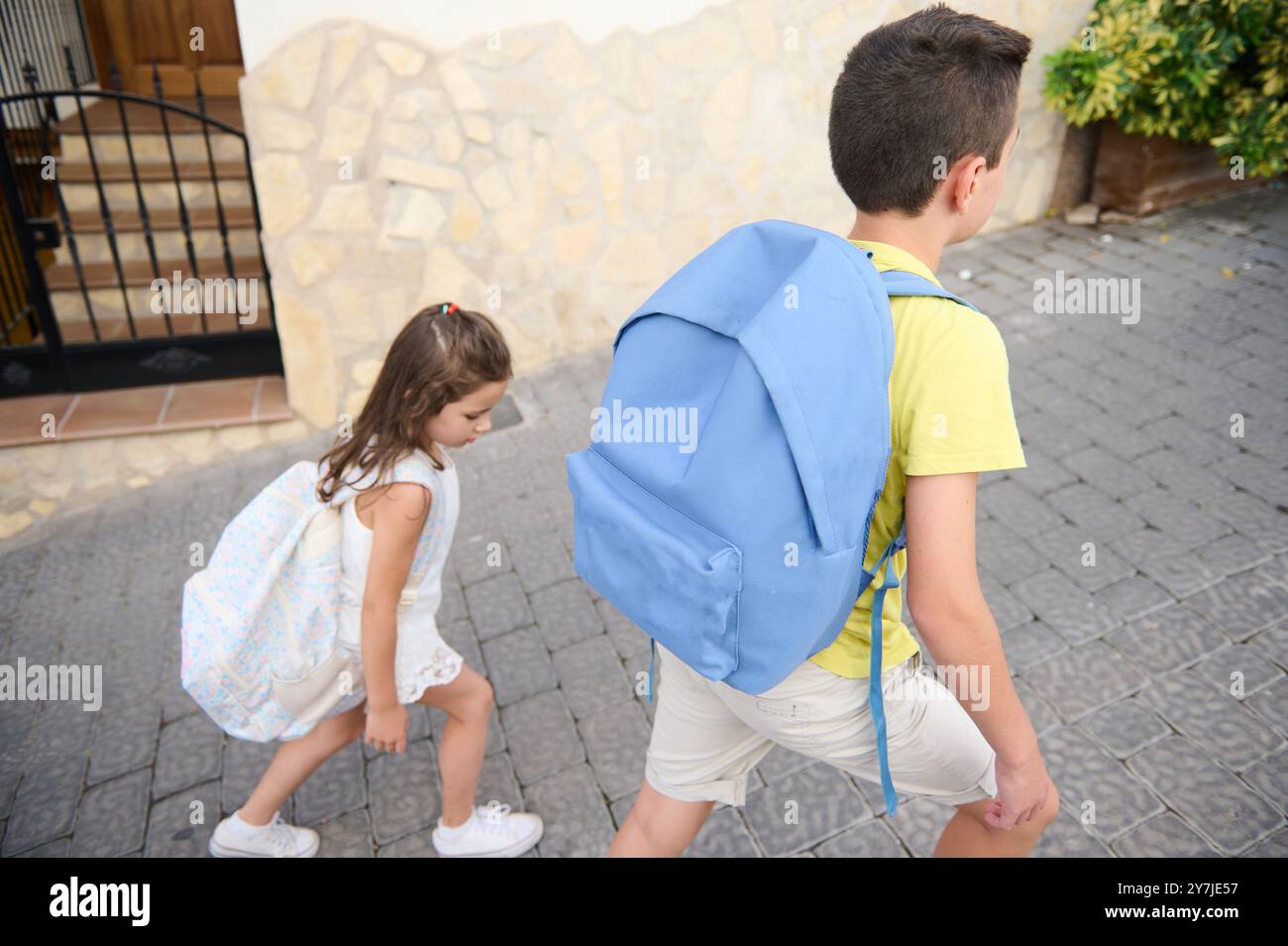 Two children with backpacks walking on a paved street on their way to ...