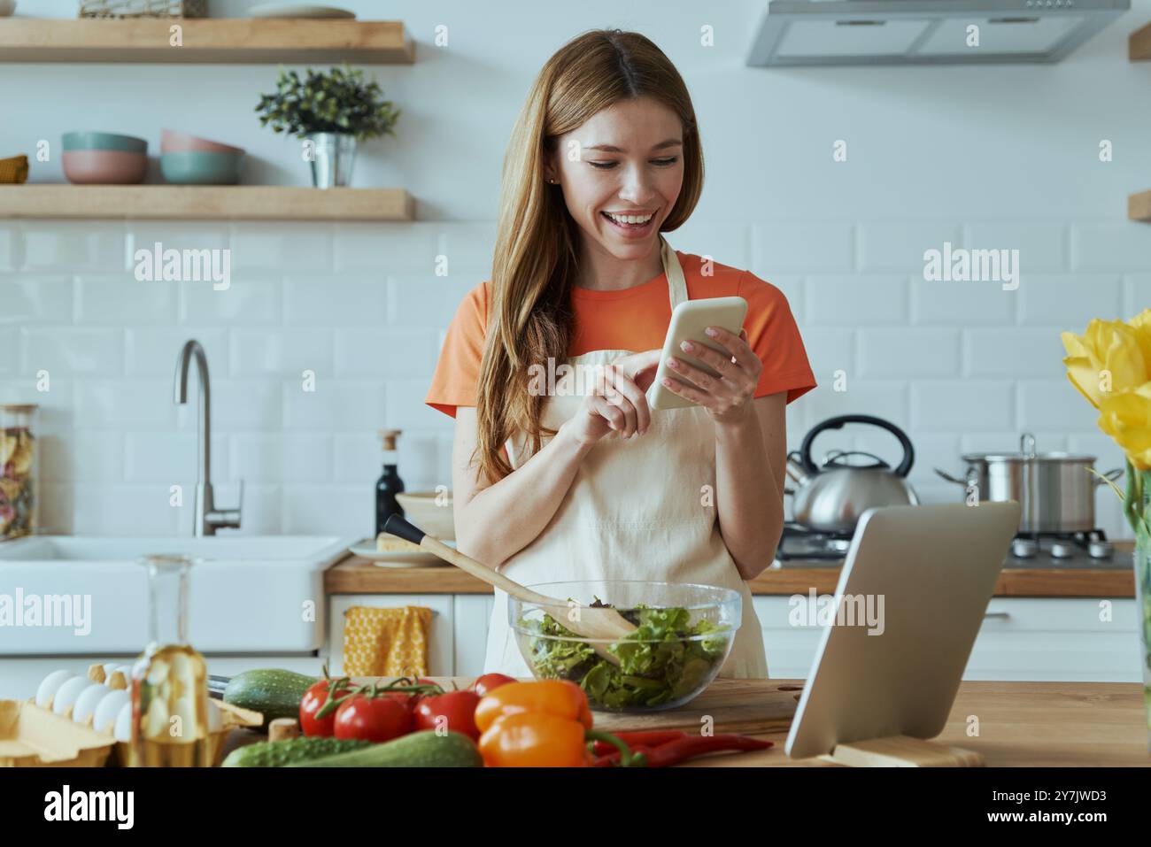 Happy young woman using technologies while cooking at the domestic ...