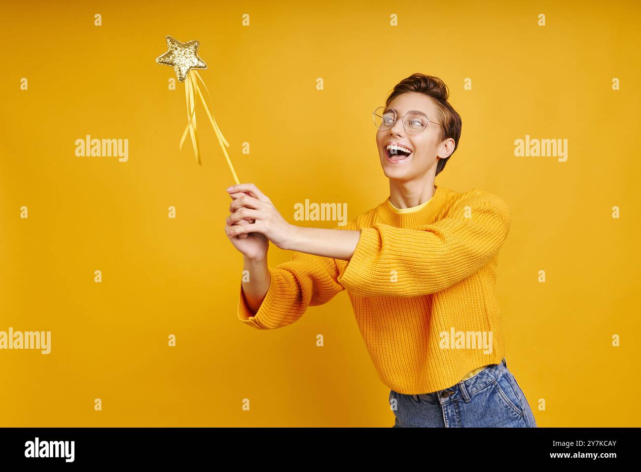 Playful young woman holding magic wand while standing against yellow ...
