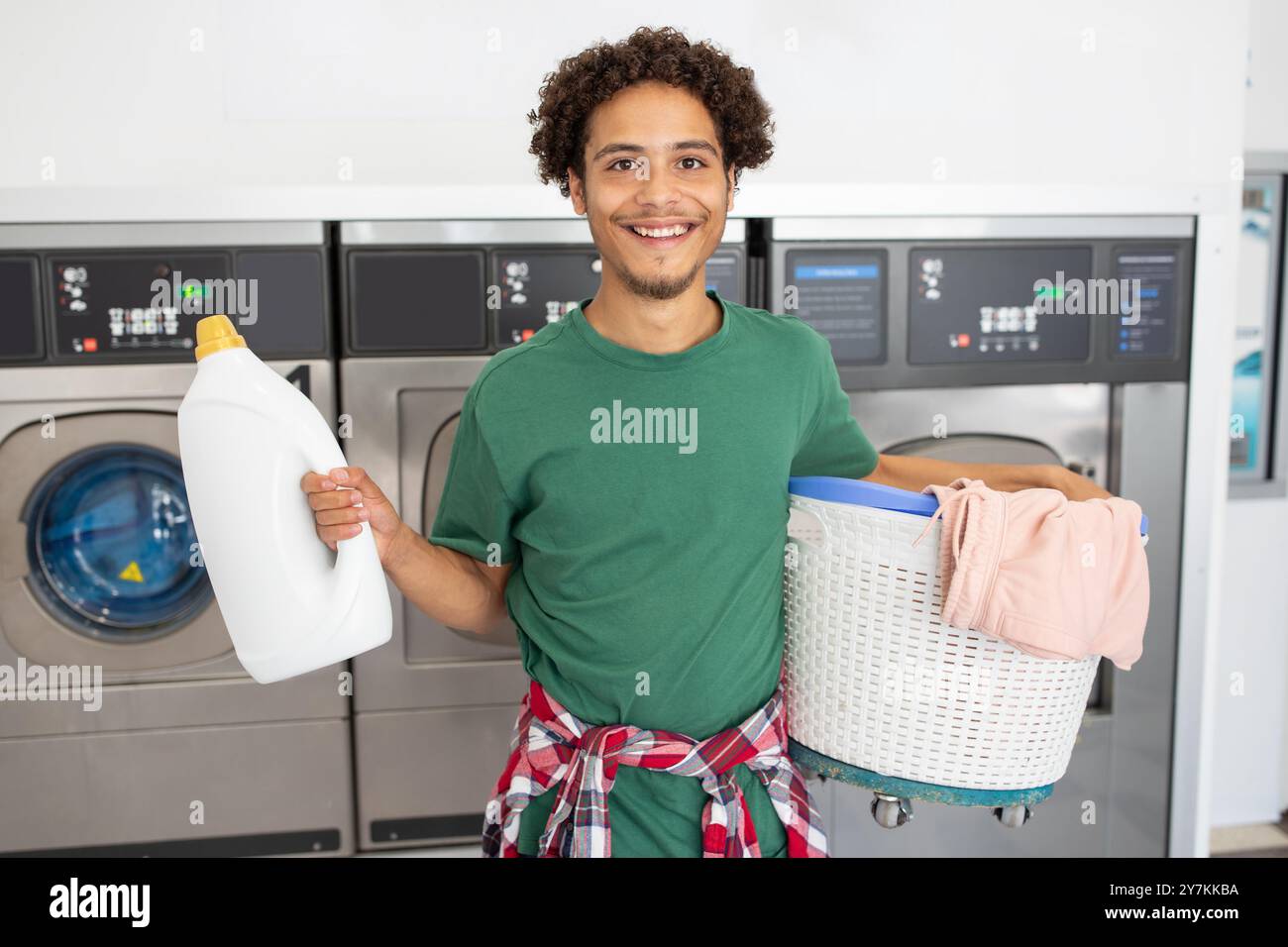 Smiling young man carrying laundry basket and detergent in laundromat ...