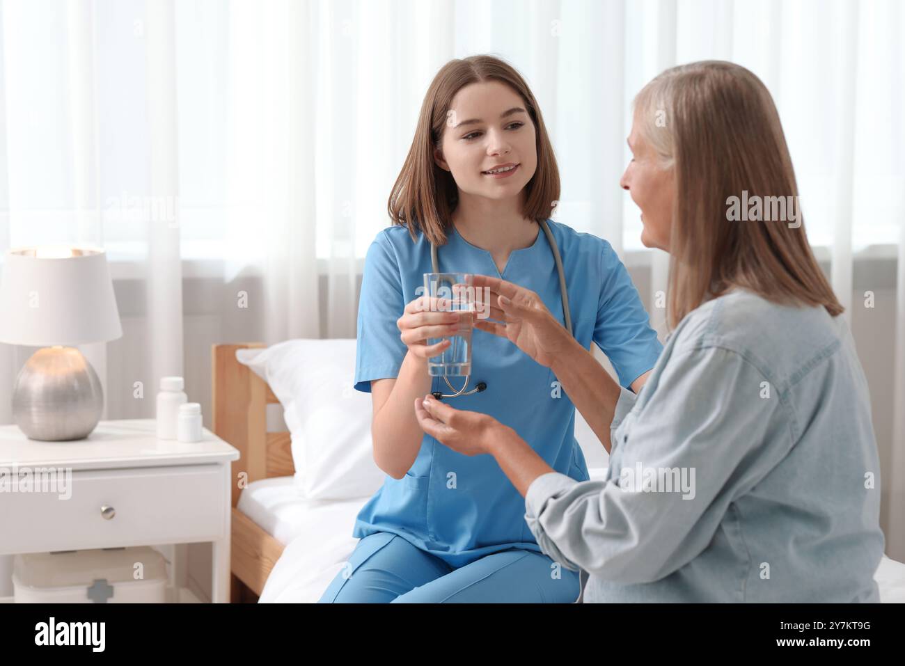 Young healthcare worker giving glass of water to senior patient on bed ...