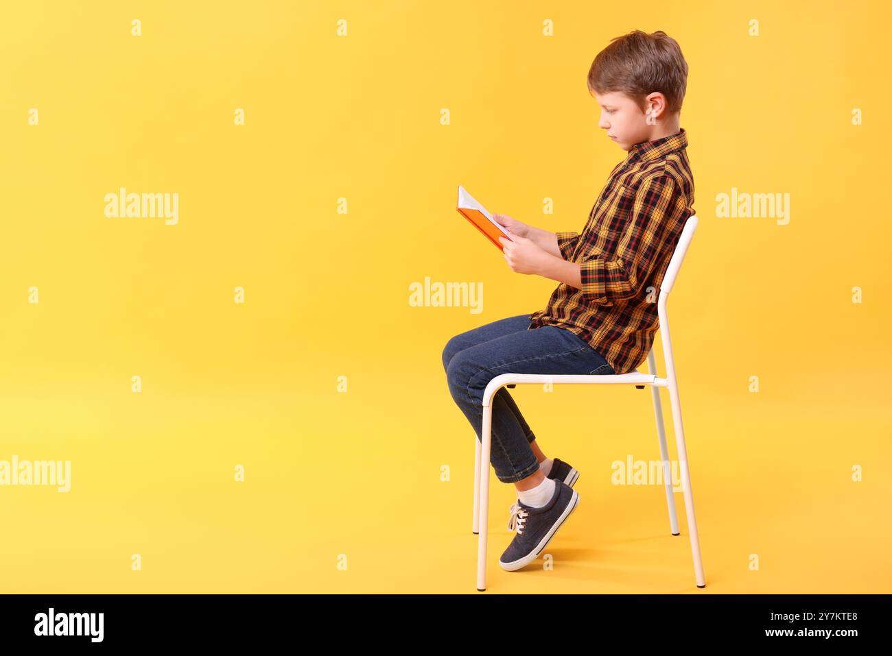 Boy with correct posture reading book on chair against yellow ...